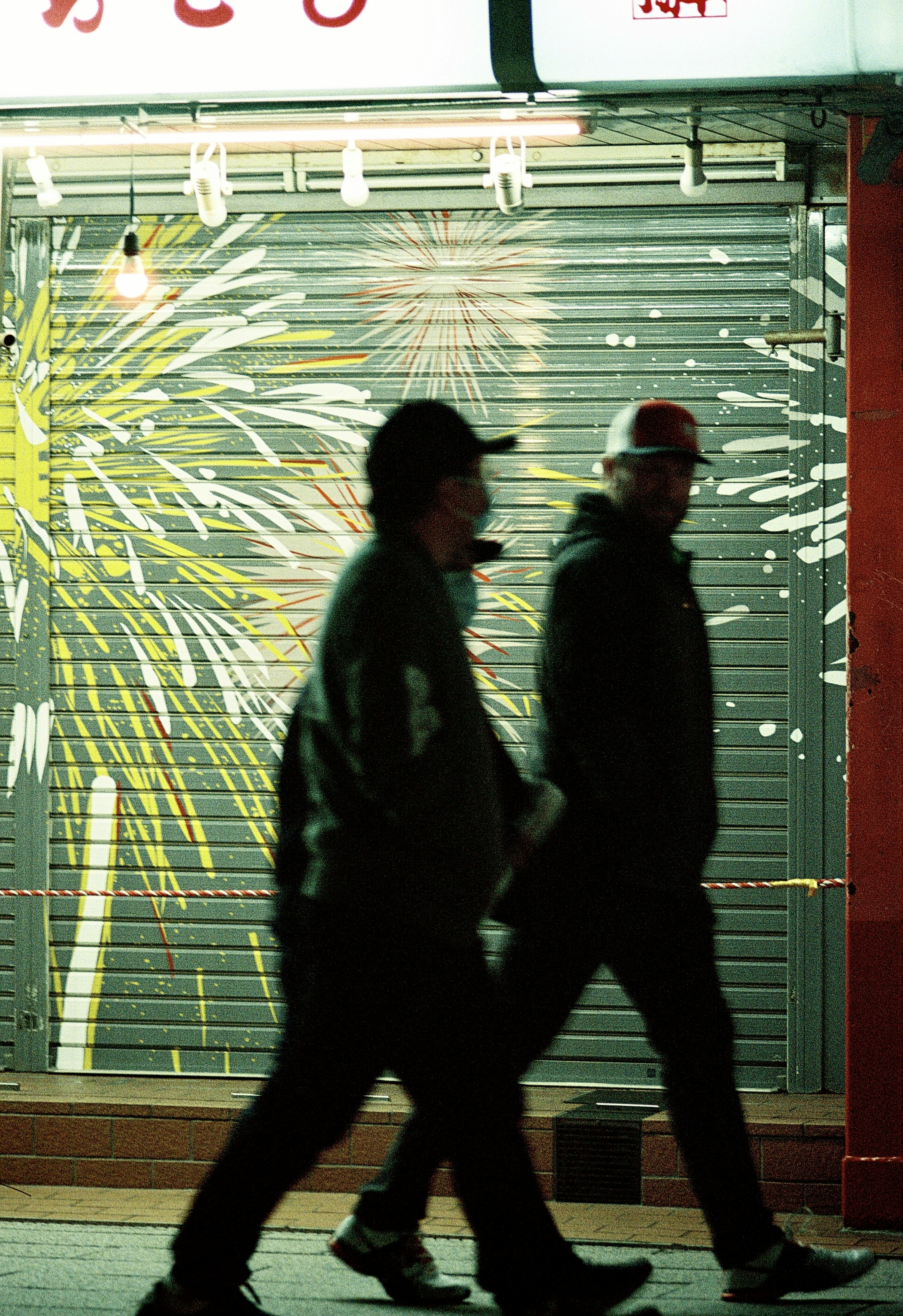 Two figures stroll past a colorful mural featuring fireworks on a shuttered storefront. The scene captures the essence of urban nightlife.