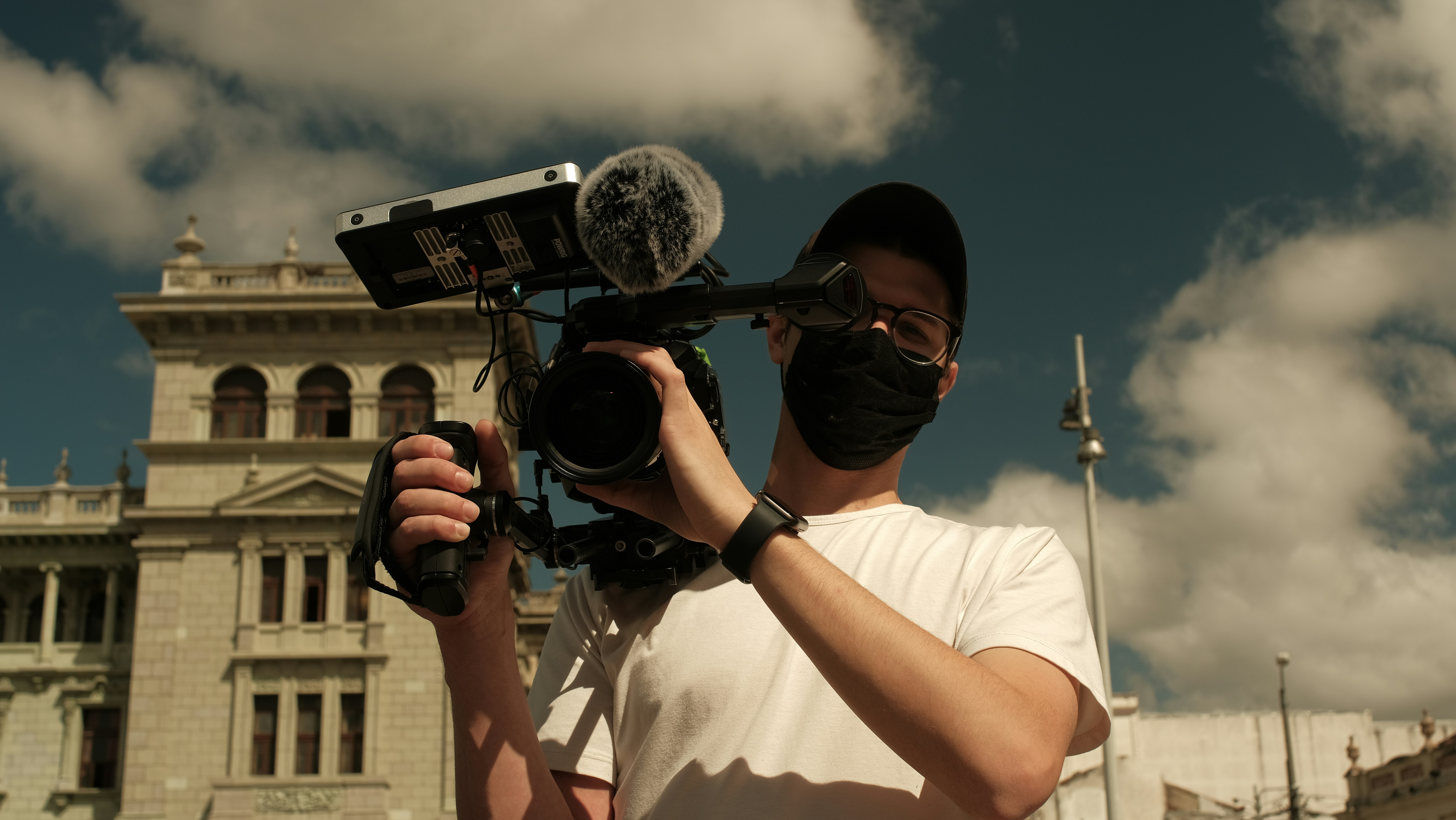 A videographer equipped with a camera and microphone captures the vibrant atmosphere in an urban setting, framed by historic architecture.