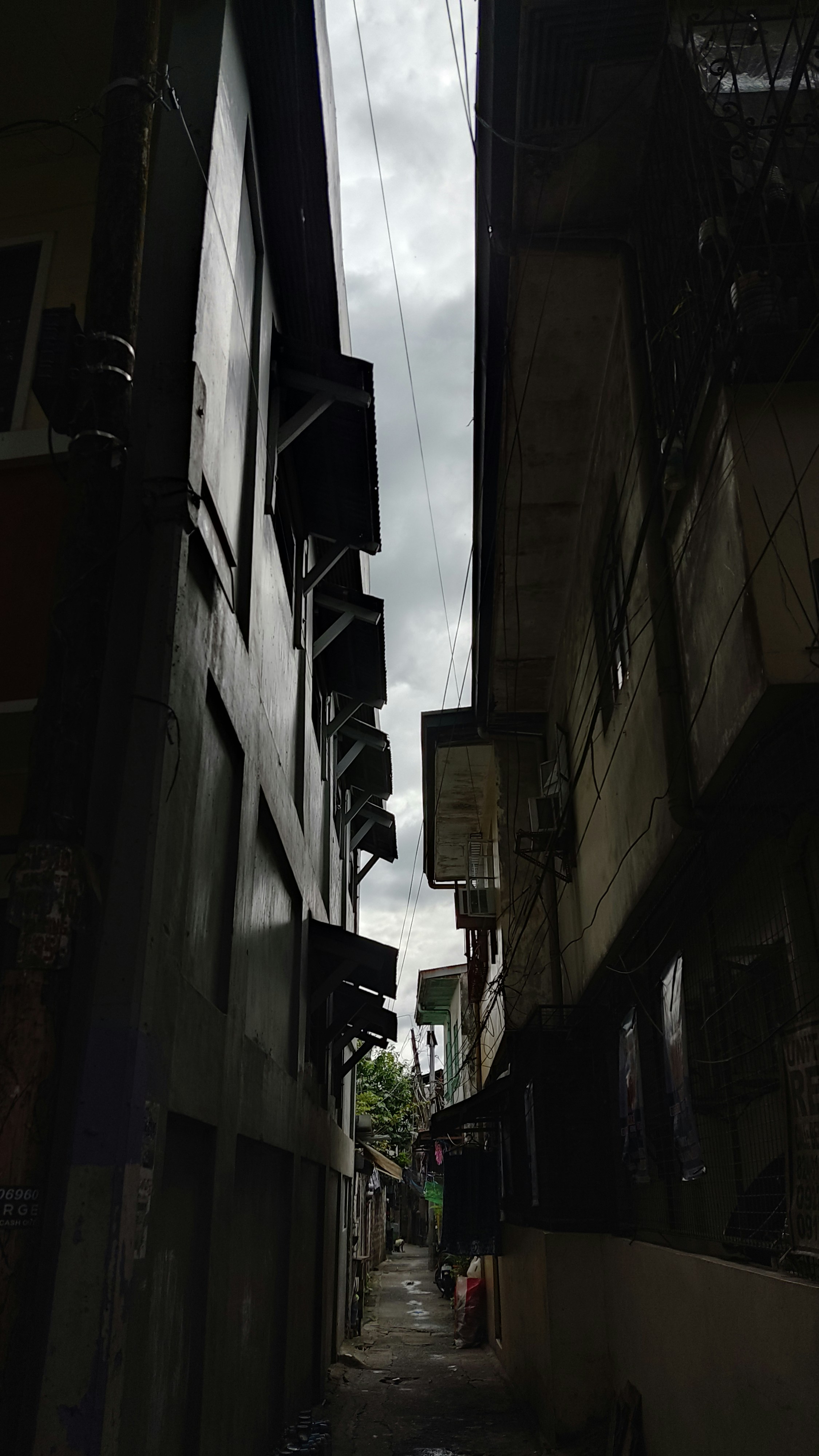 Narrow alleyway flanked by tall buildings under a cloudy sky, revealing hints of urban life. Clothes hang to dry, adding a touch of color to the muted surroundings.