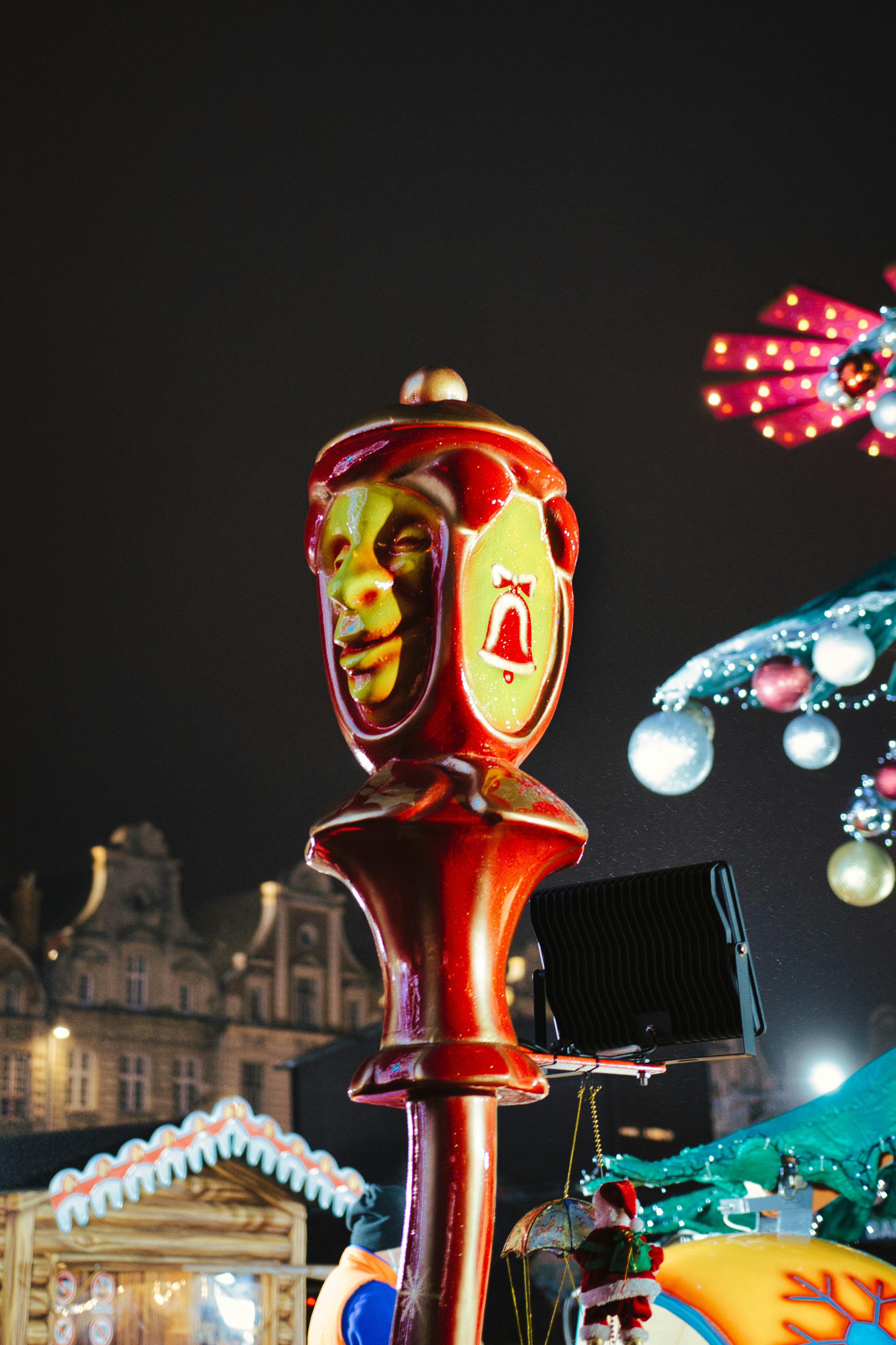 A vibrant red pole featuring a whimsical face and bell, adorned with colorful decorations in a festive setting. The background showcases a charming holiday market atmosphere.