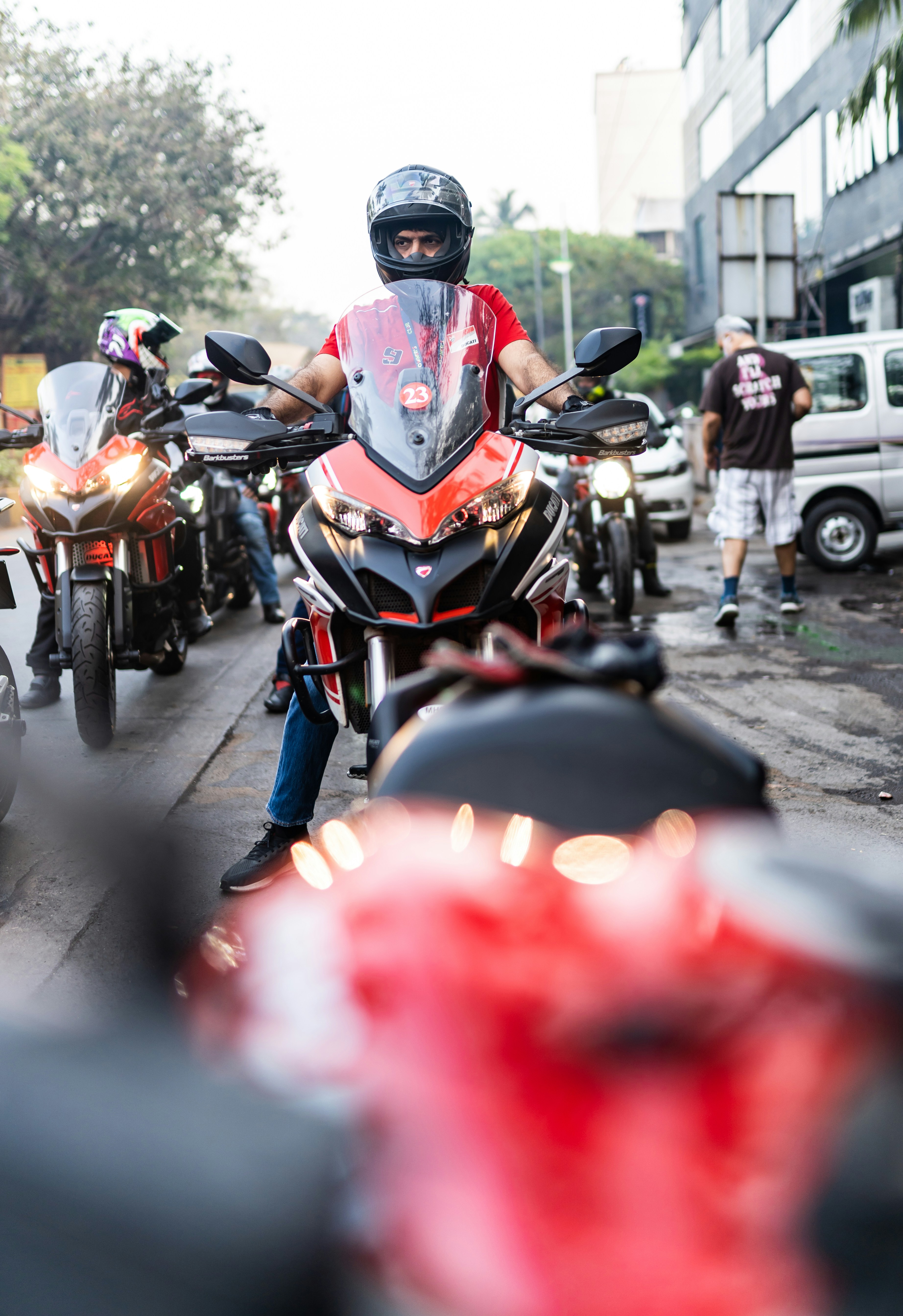 Motorcyclist in a vibrant red outfit sits confidently on a motorcycle, surrounded by a bustling urban environment. The focus highlights the rider's determination amidst the chaos. 