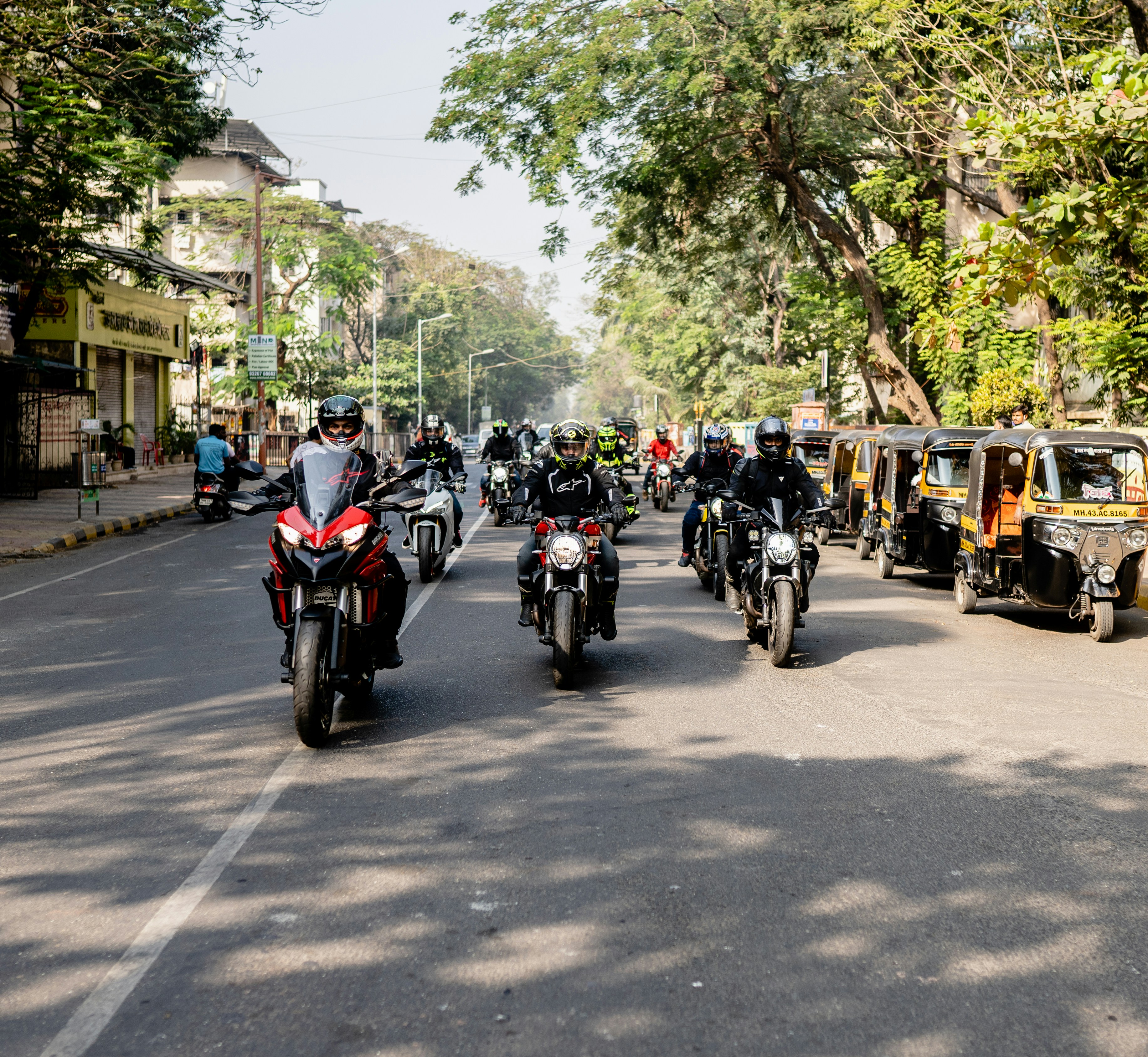 Motorcyclists navigating a bustling city street, flanked by colorful auto rickshaws and lush greenery. The scene captures the vibrant energy of urban life.