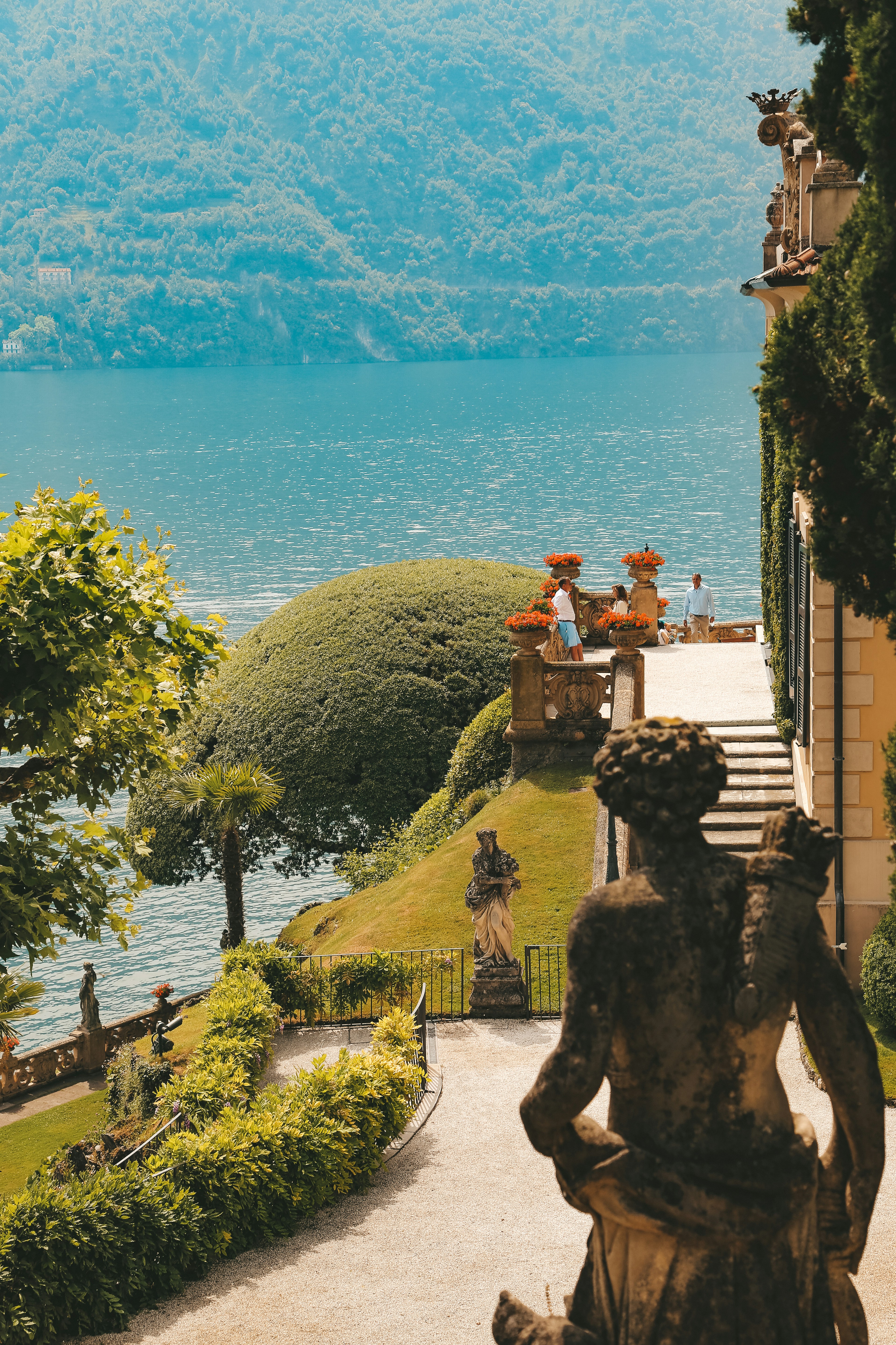 Elegant garden path leading to a serene lake, framed by lush greenery and classical sculptures. A couple enjoys the view from a terrace.