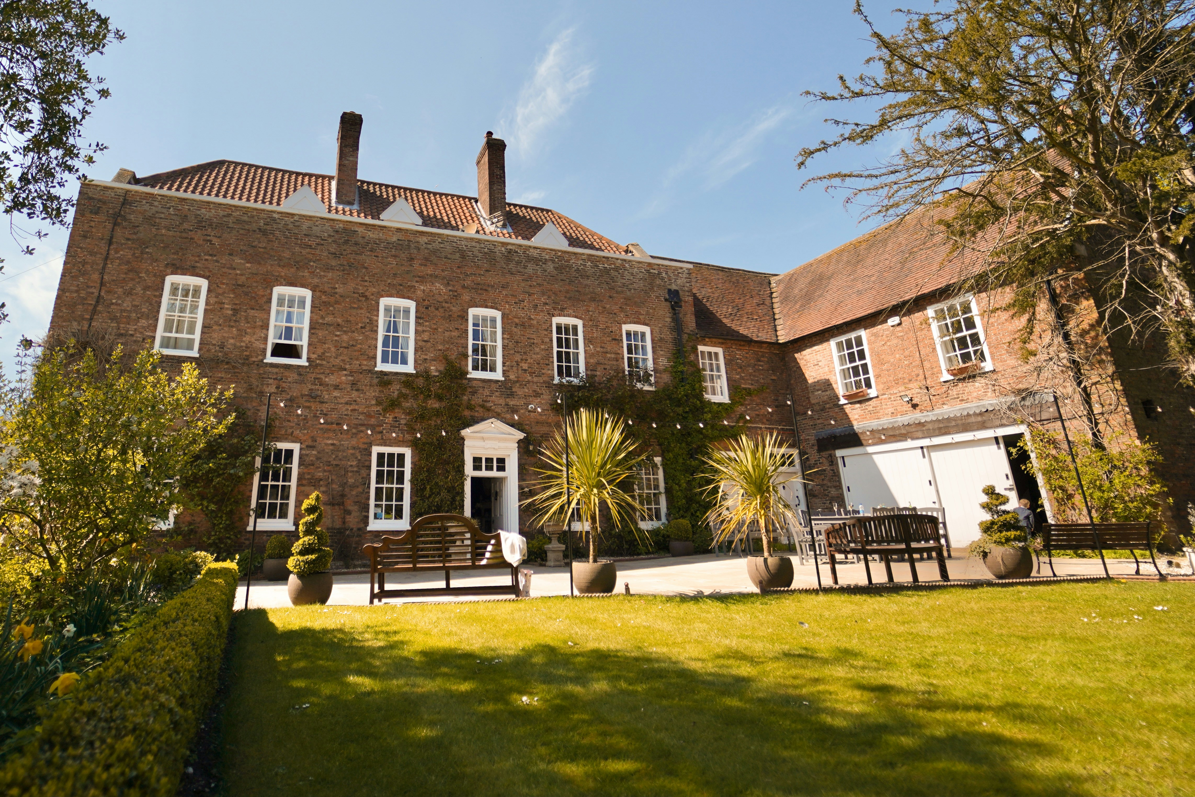Elegant brick building with large windows surrounded by lush greenery and garden furniture. The scene evokes a sense of tranquility and outdoor leisure.