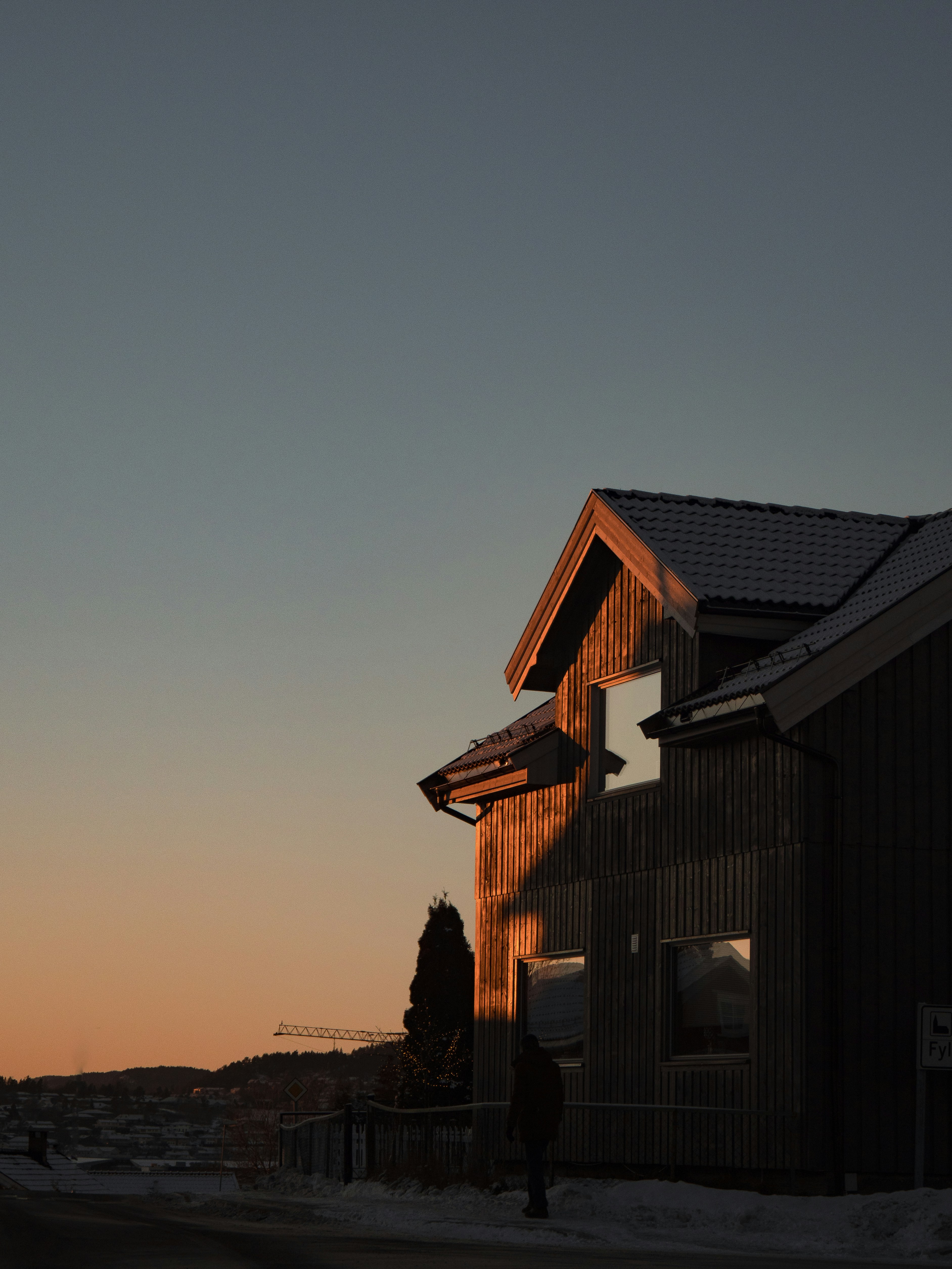 Sunset casts warm light on a contemporary wooden house, silhouetted against a fading sky. A figure stands nearby, adding a sense of scale.