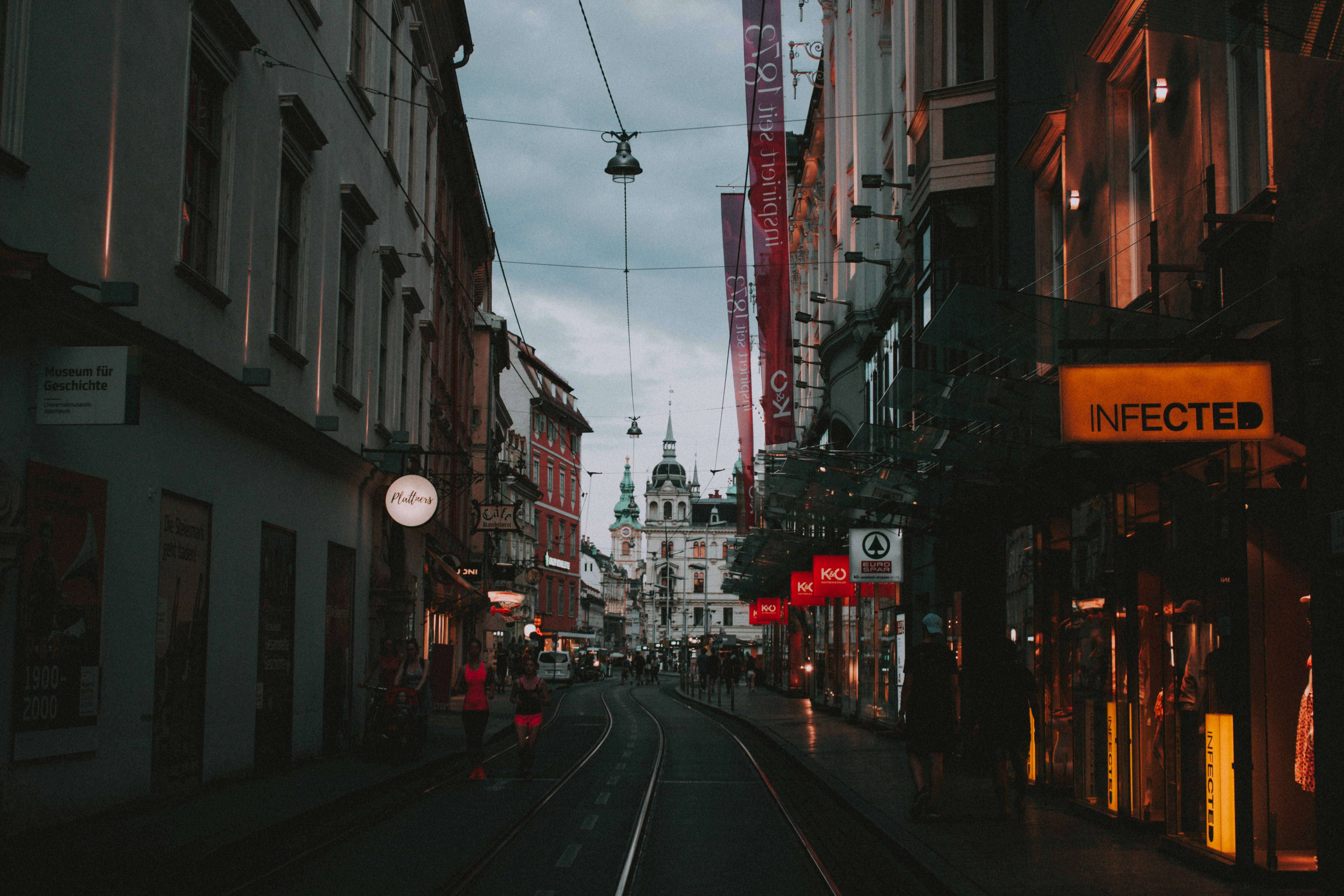 A dimly lit city street at dusk, with tram tracks leading toward a distant illuminated church.