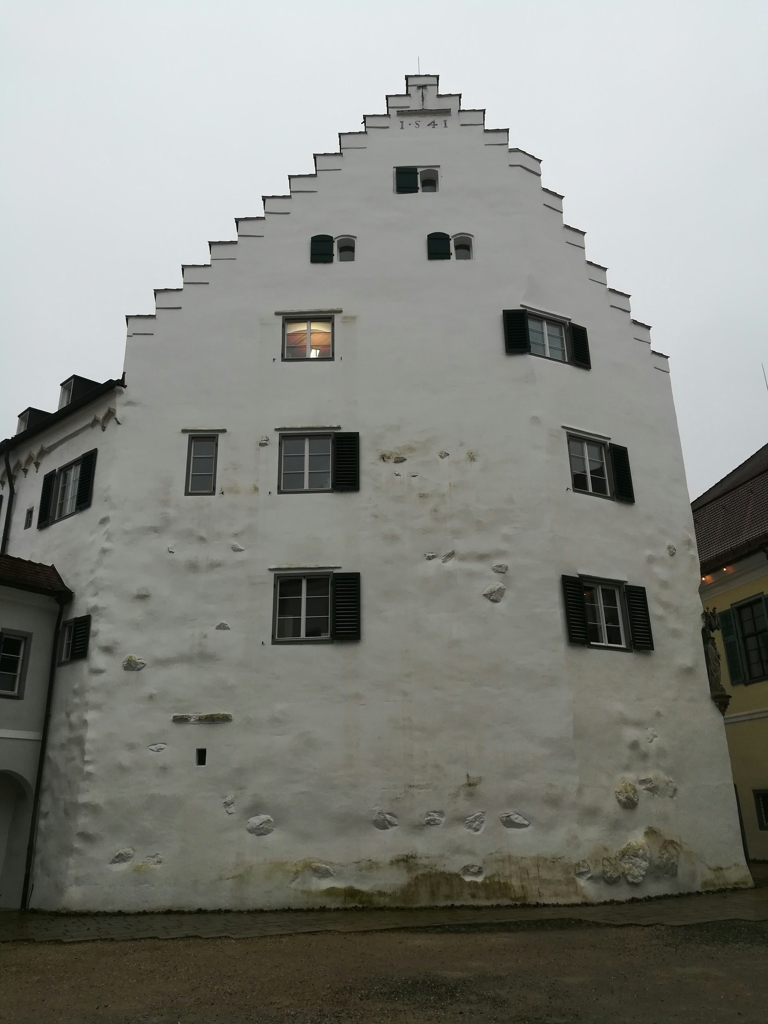 Tall white building with a stepped gable and dark shutters rises against an overcast sky. A single interior window glows warmly, drawing the eye.