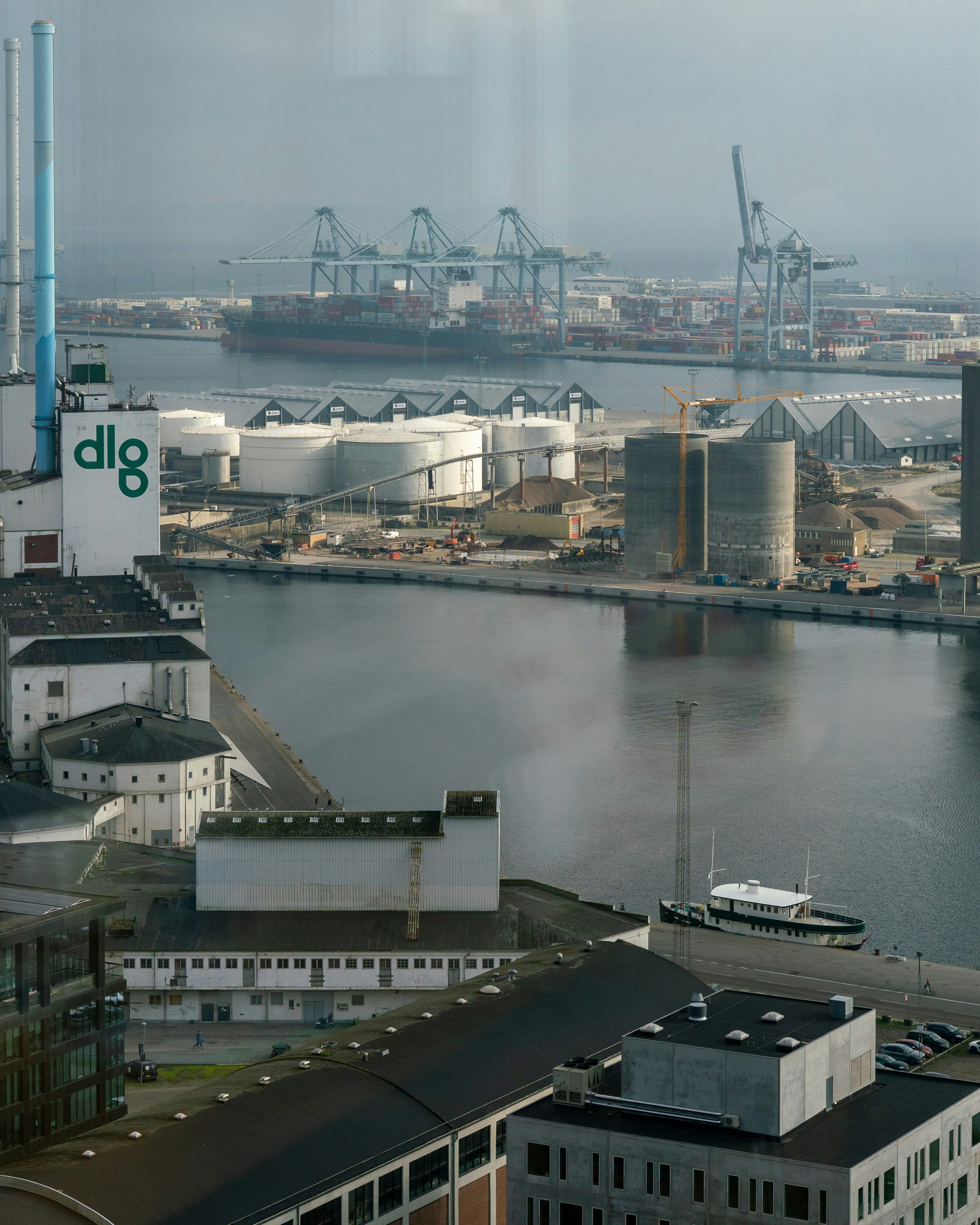 A panoramic view of a busy industrial port featuring storage tanks, cargo ships, and cranes against a cloudy sky.