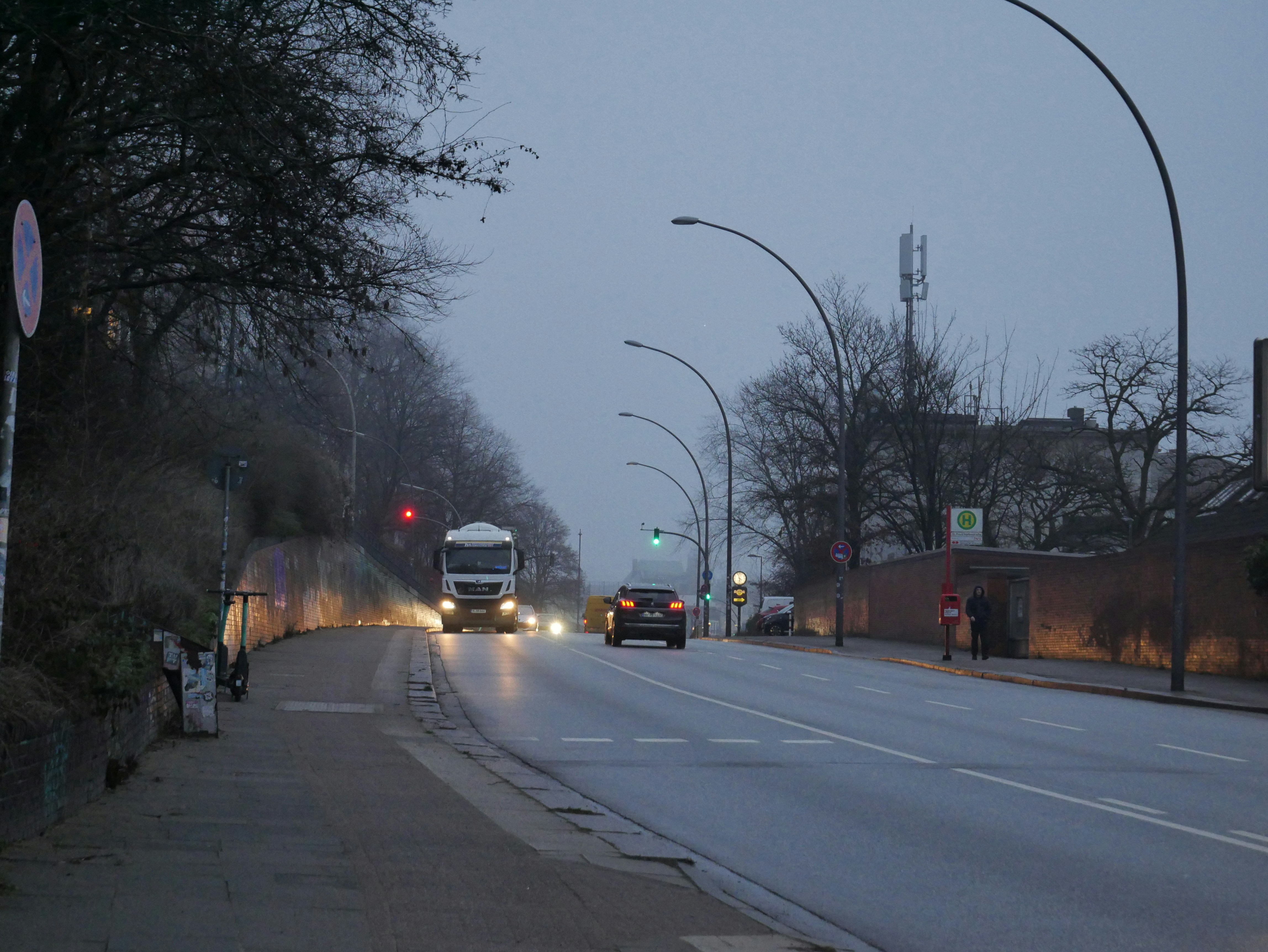 A foggy urban street at dawn, featuring vehicles approaching traffic lights and silhouetted trees along the roadside.