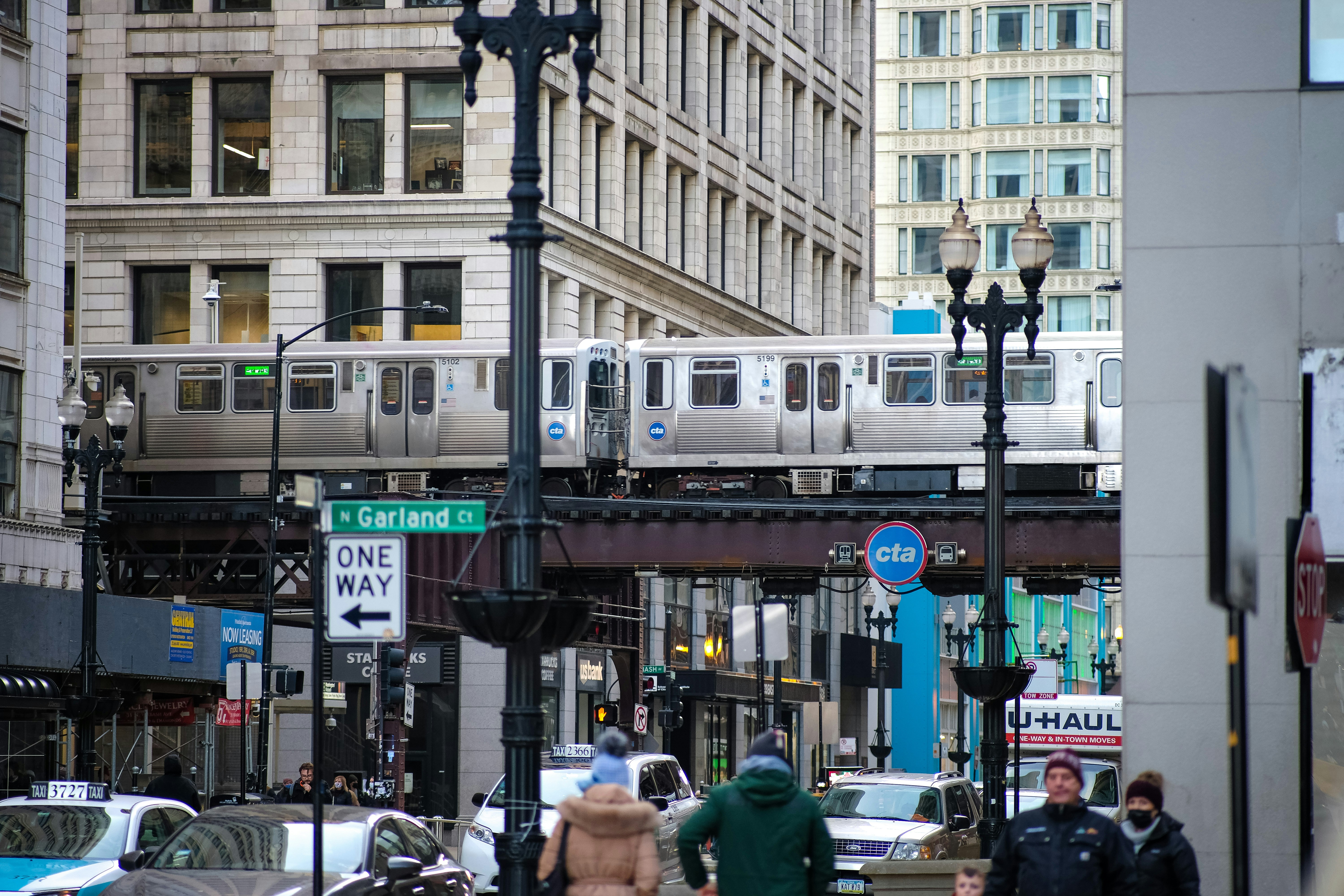Silver train gliding above bustling city street, framed by historic architecture and modern buildings. Traffic and pedestrians create a dynamic urban scene.