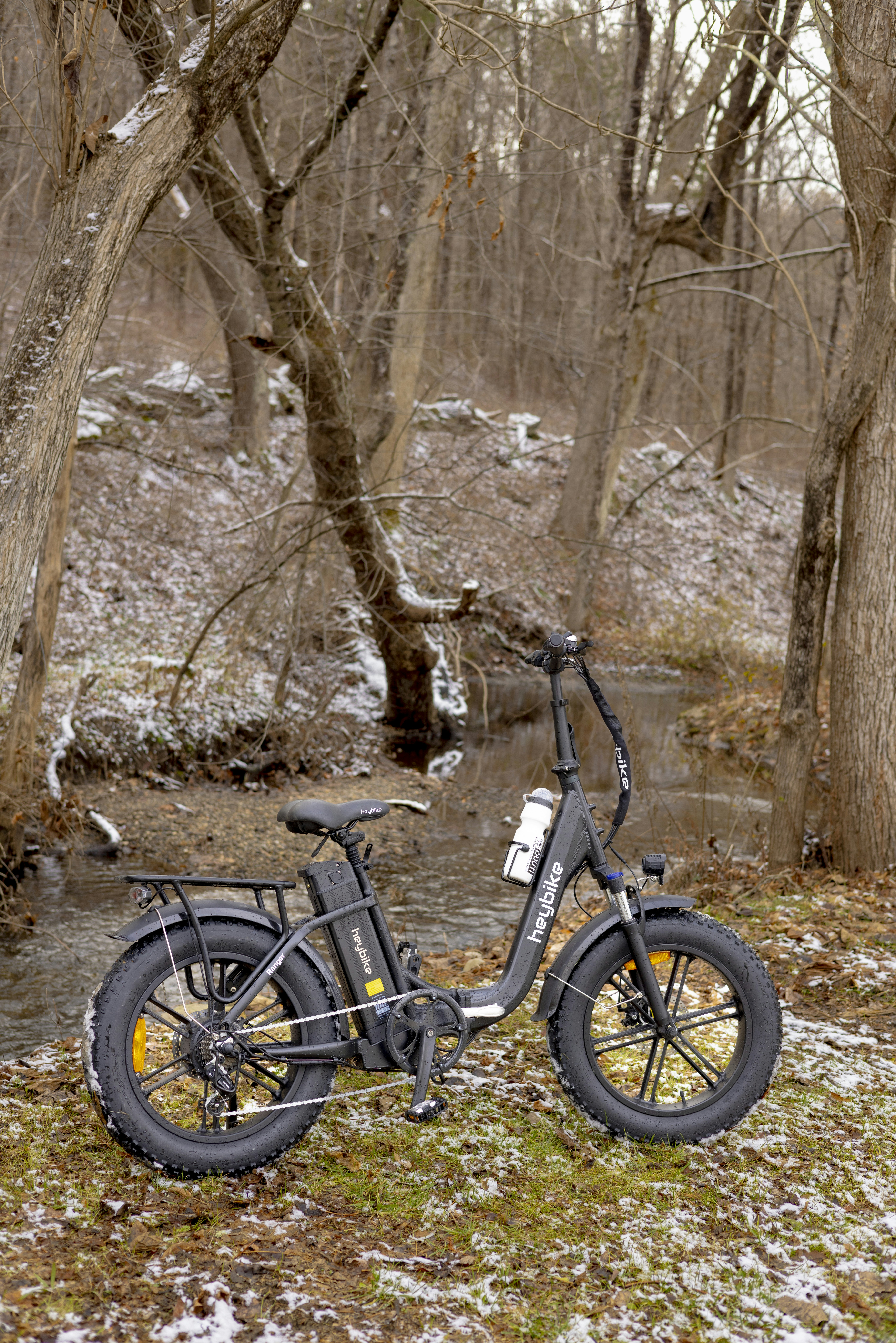 Fat tire e-bike parked beside a gently flowing stream in a winter landscape, surrounded by leafless trees and a dusting of snow.
