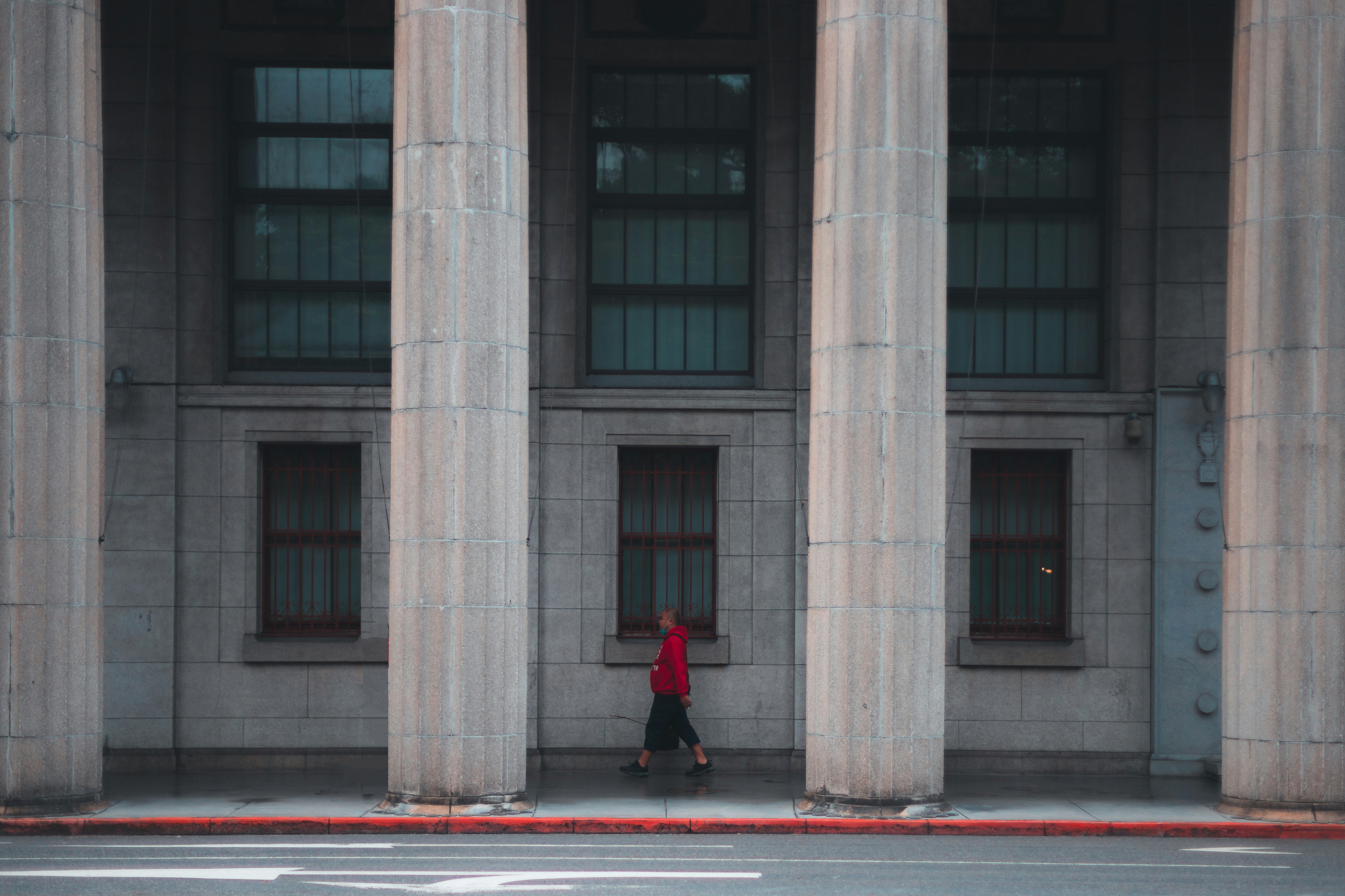 A figure in a red coat walks along a stone colonnade, framed by towering columns and large windows, evoking a sense of isolation in an urban landscape.