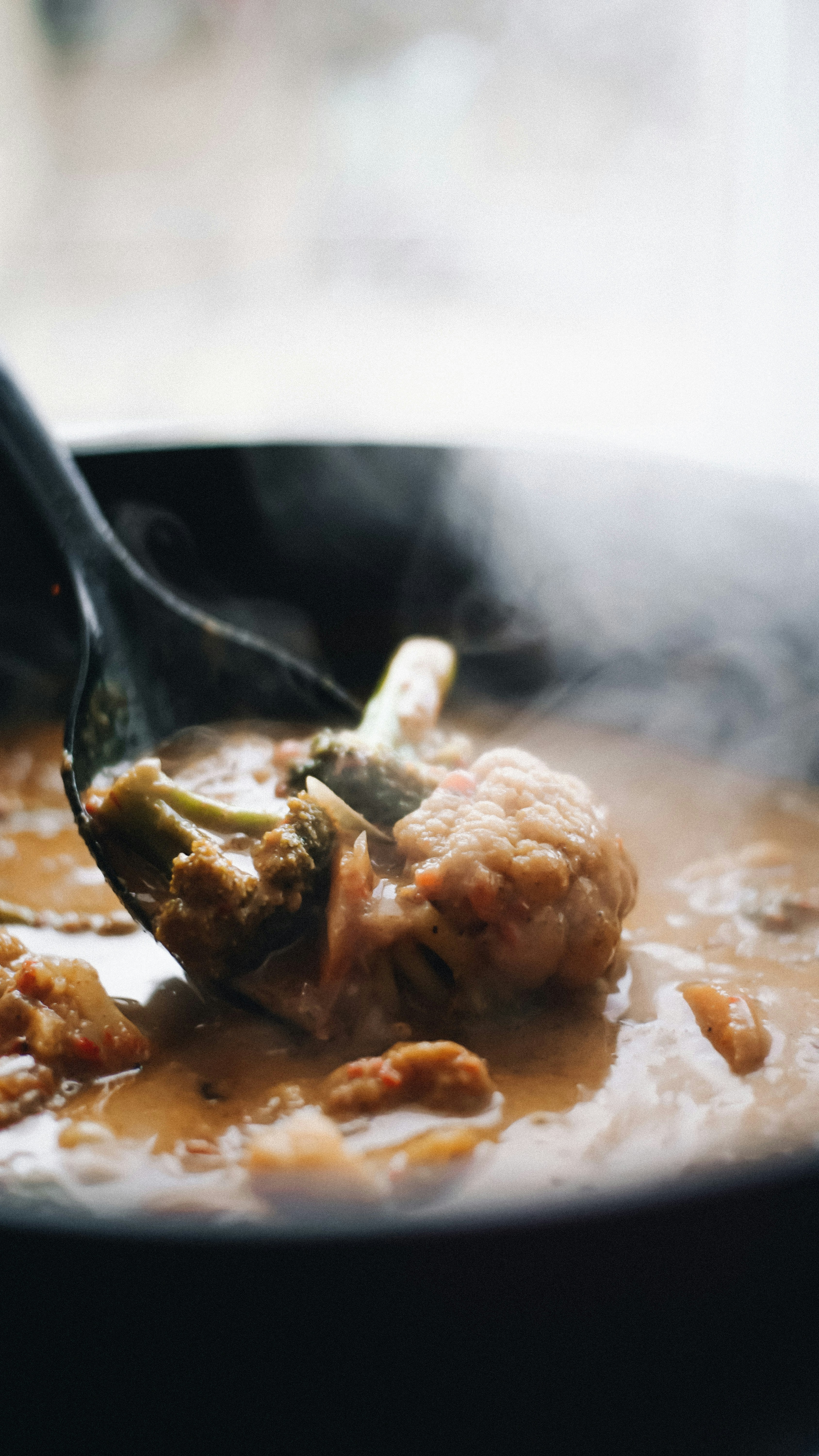 Steaming bowl of savory soup with broccoli and cauliflower being served with a black ladle. The warmth and richness of the dish are highlighted by the gentle steam rising.