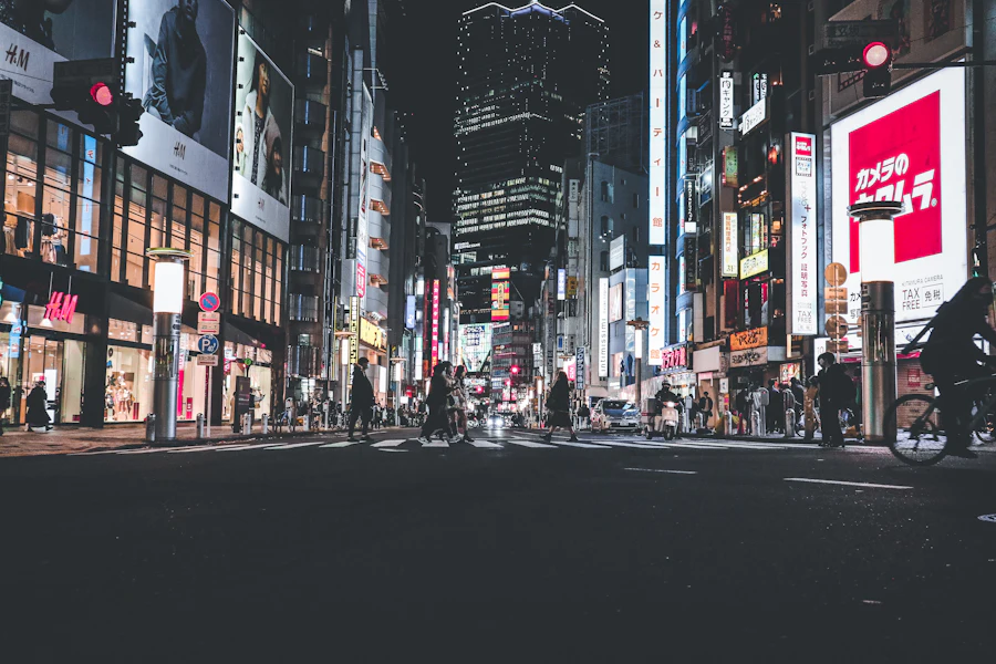Busy city street at night with pedestrians crossing between bright shop signs and tall illuminated buildings. A cyclist blurs past on the right as traffic lights glow over the intersection.