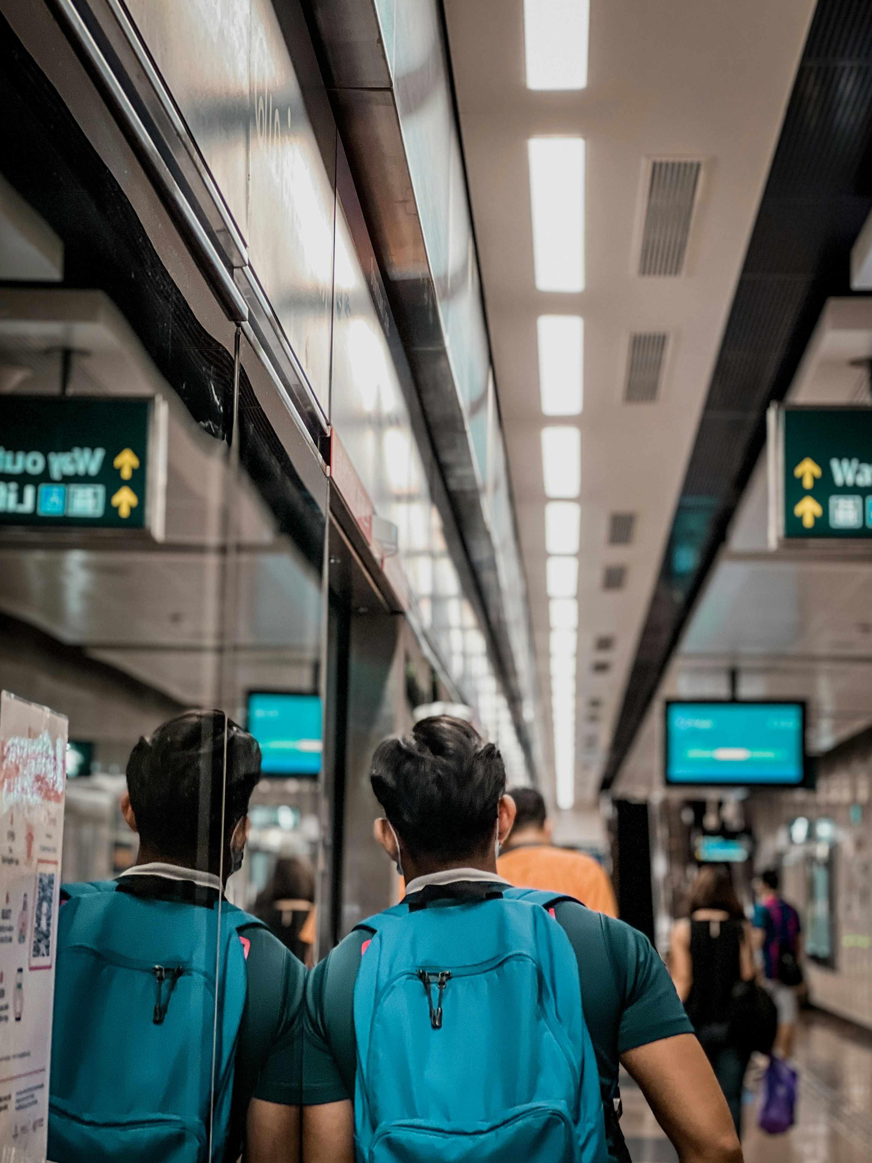 A man with a turquoise backpack stands in front of a reflective surface in a subway station, capturing the essence of urban travel. The scene is framed by overhead lights and directional signs.