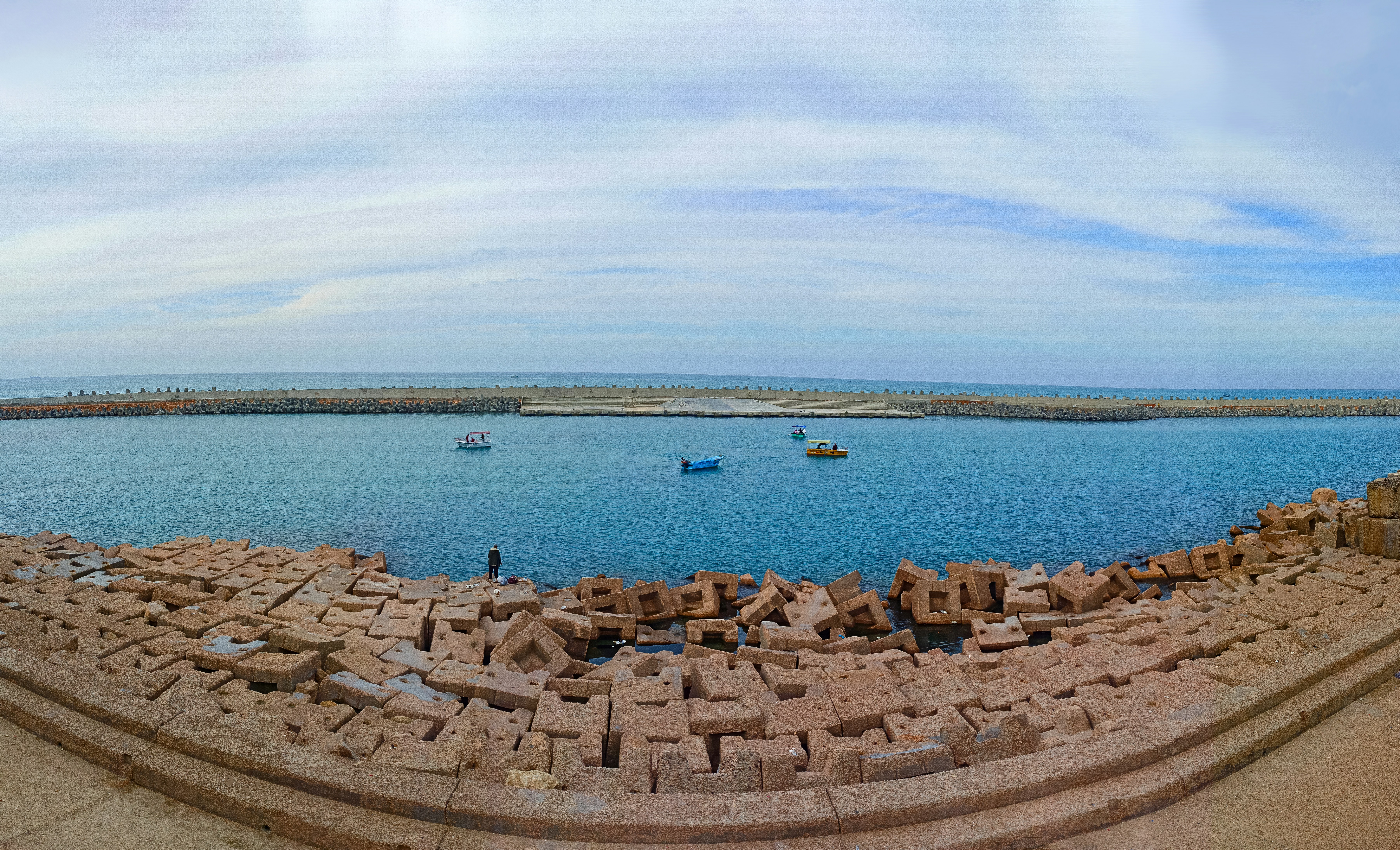 Calm blue waters with boats drift near a historic stone embankment under a cloudy sky.