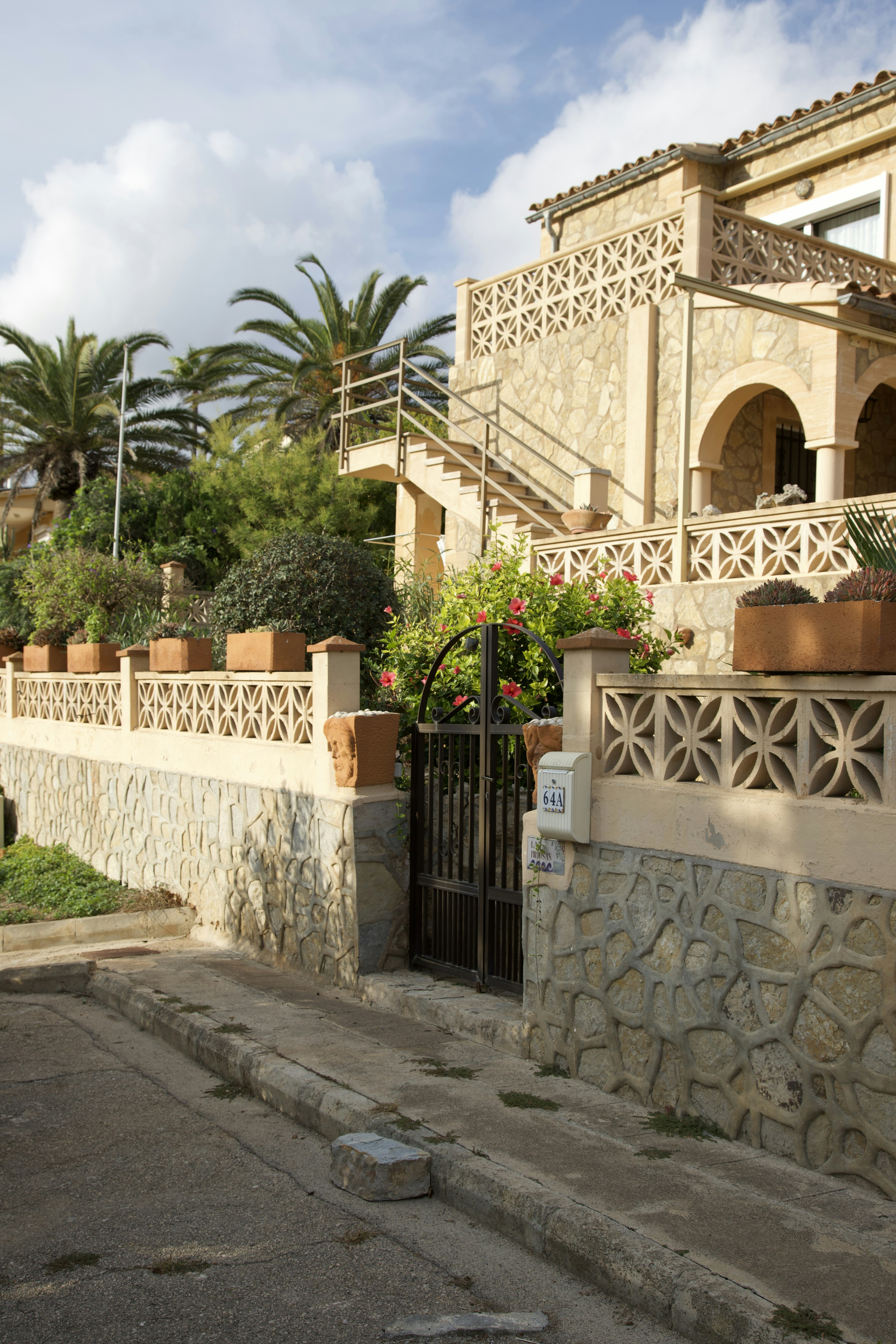Charming stone house with lush greenery and terracotta pots, nestled among palm trees under a bright sky.