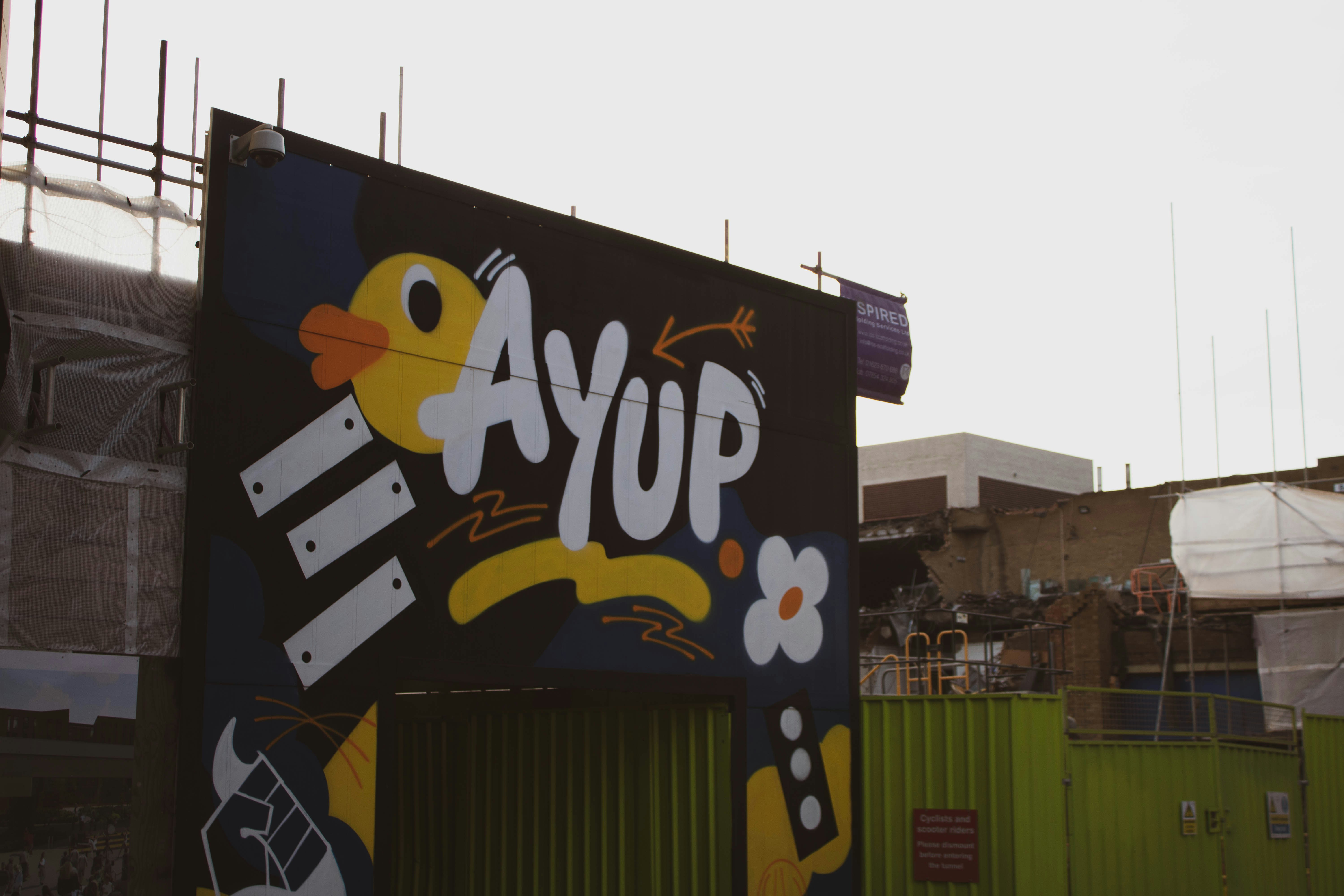 Colorful mural featuring a cartoon bird and the word 'AYUP' on a construction site.