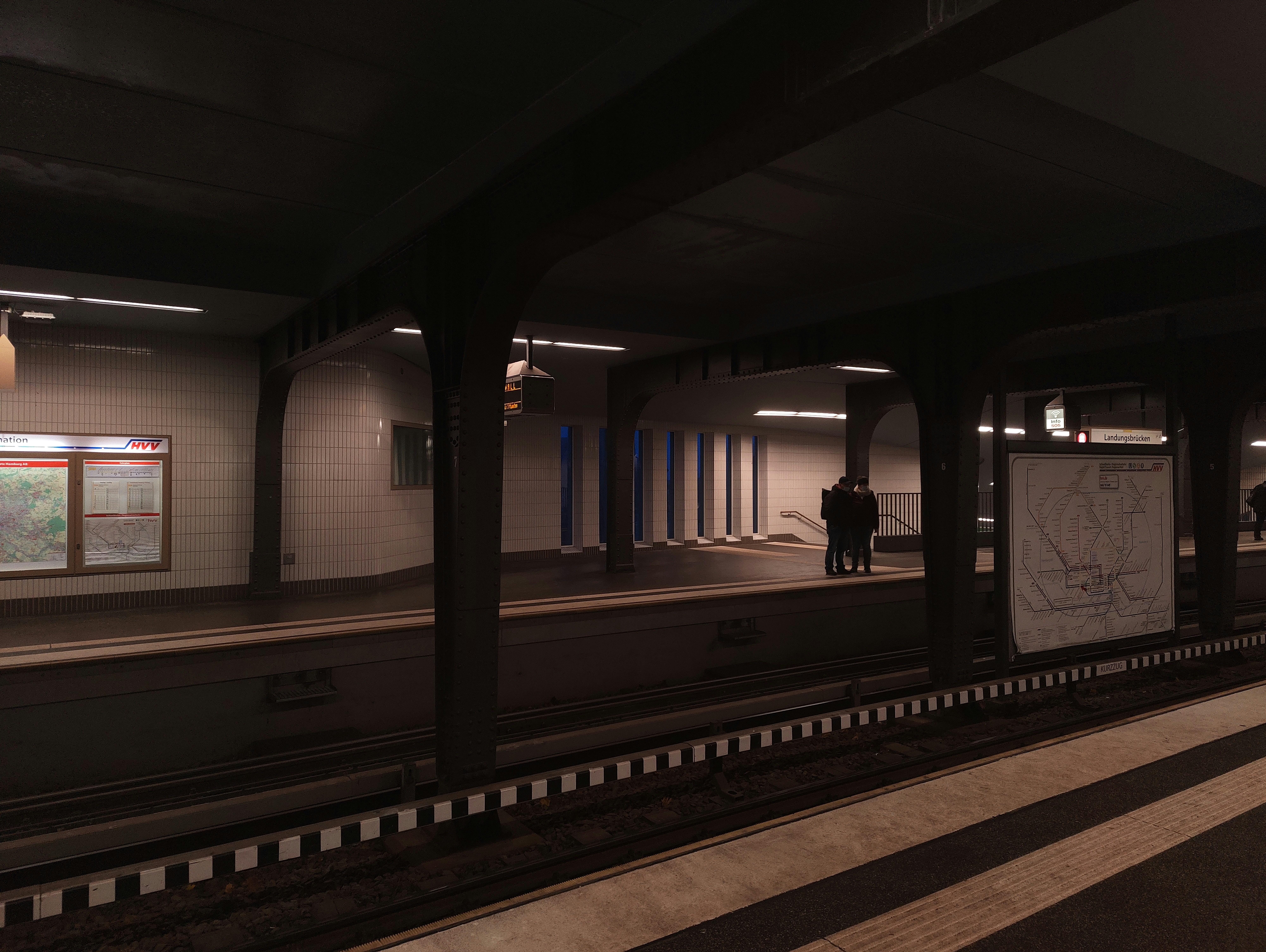 Dimly lit subway platform with tiled walls and heavy arches, featuring a map board and two figures near the far track.