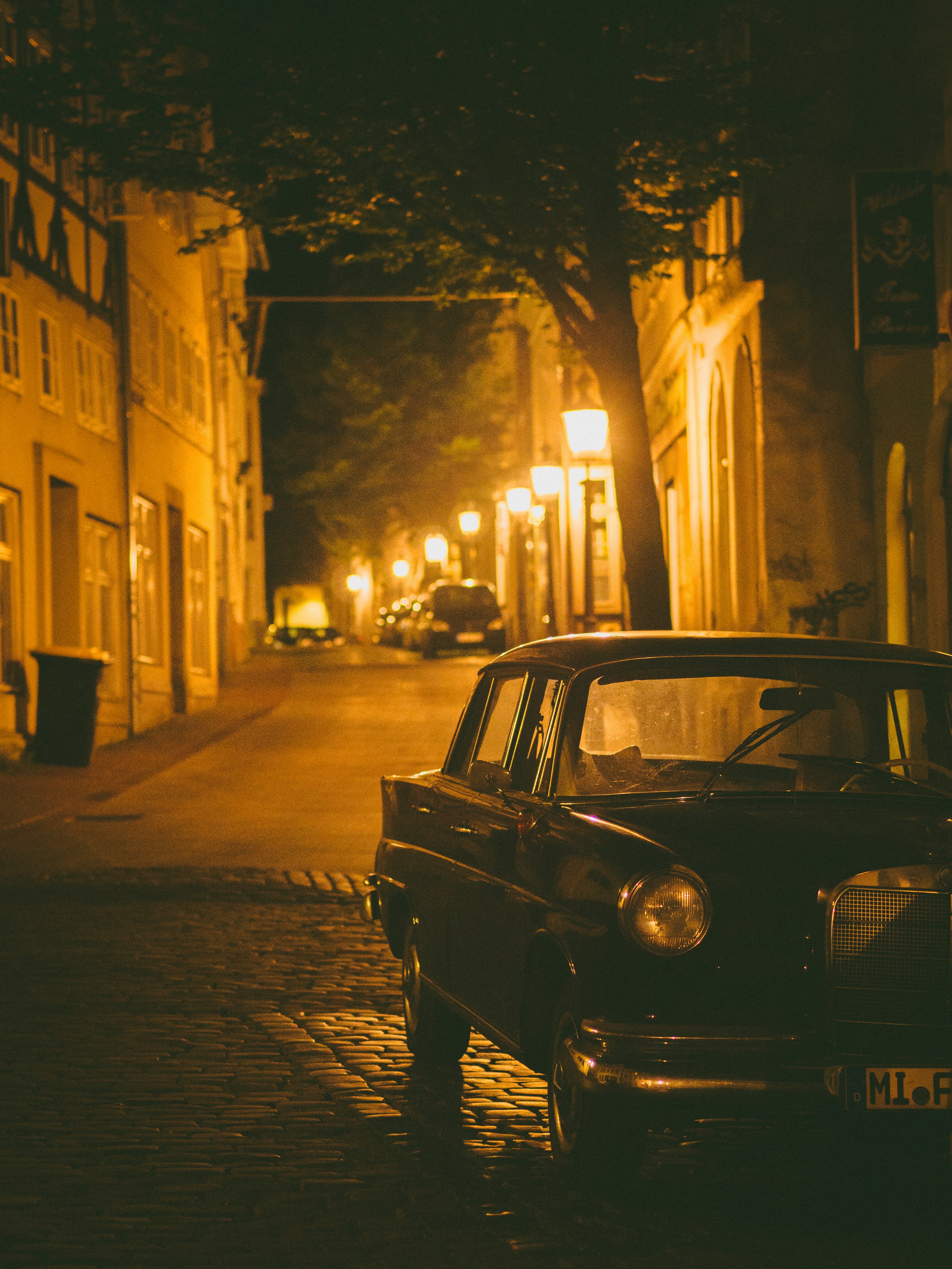 Vintage car parked on a cobblestone street illuminated by warm streetlights in a quiet nighttime setting.