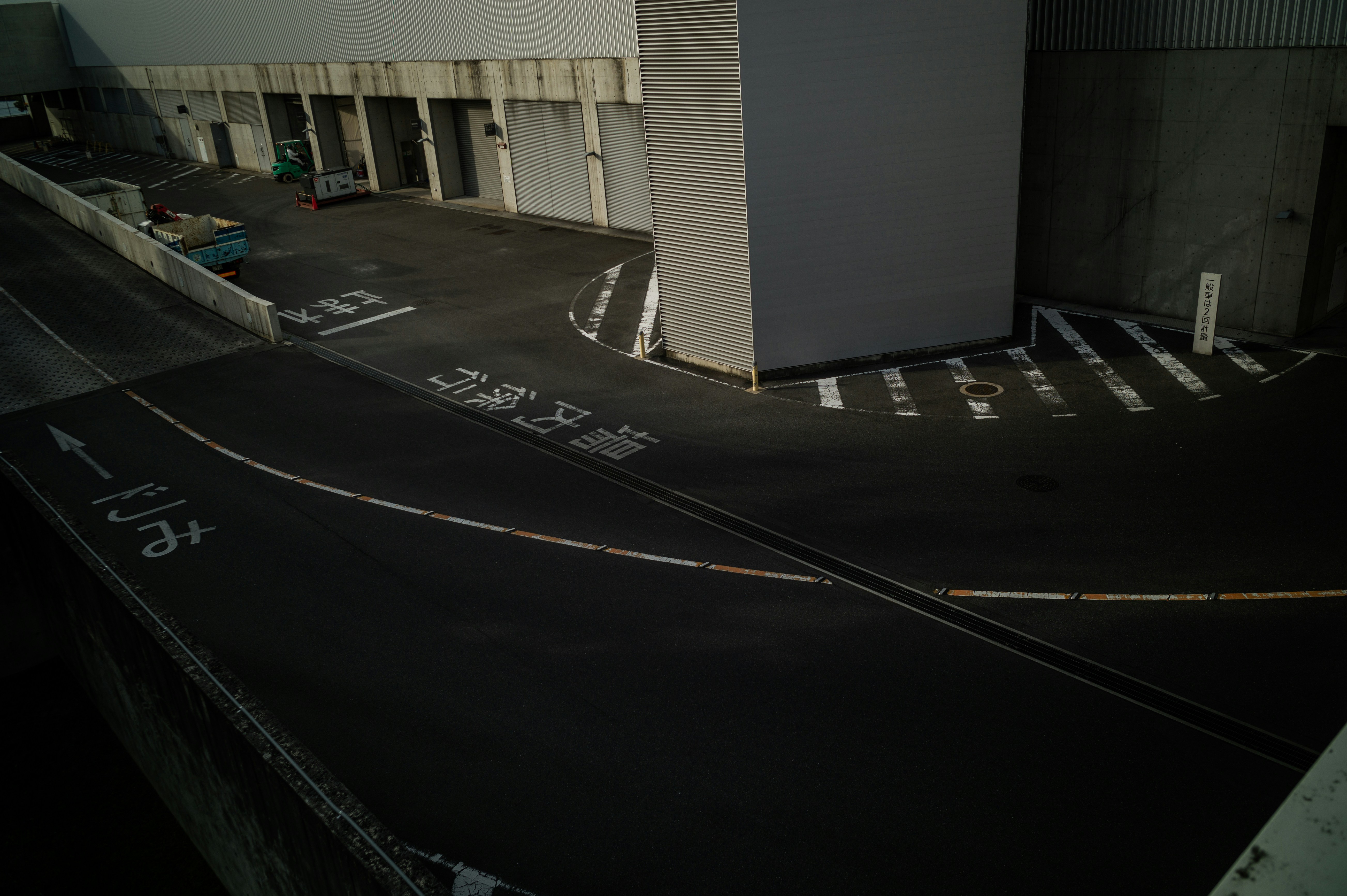 An industrial scene showcasing a winding road with directional markings and a backdrop of muted architecture. The interplay of light and shadow highlights the textures of the surfaces.