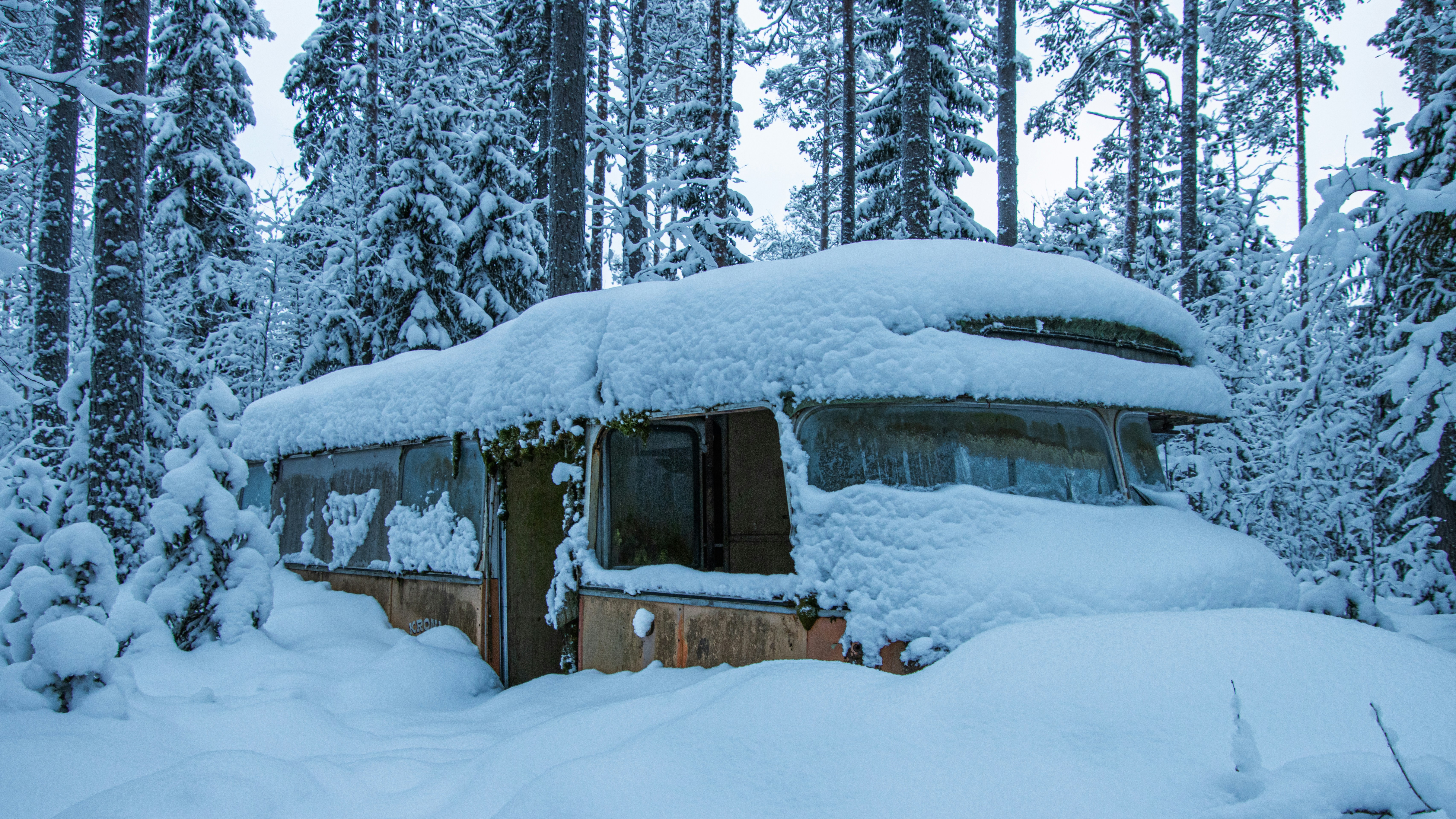 An old, snow-covered bus sits silently in a winter forest, surrounded by tall trees blanketed in white. The scene evokes a sense of nostalgia and solitude.