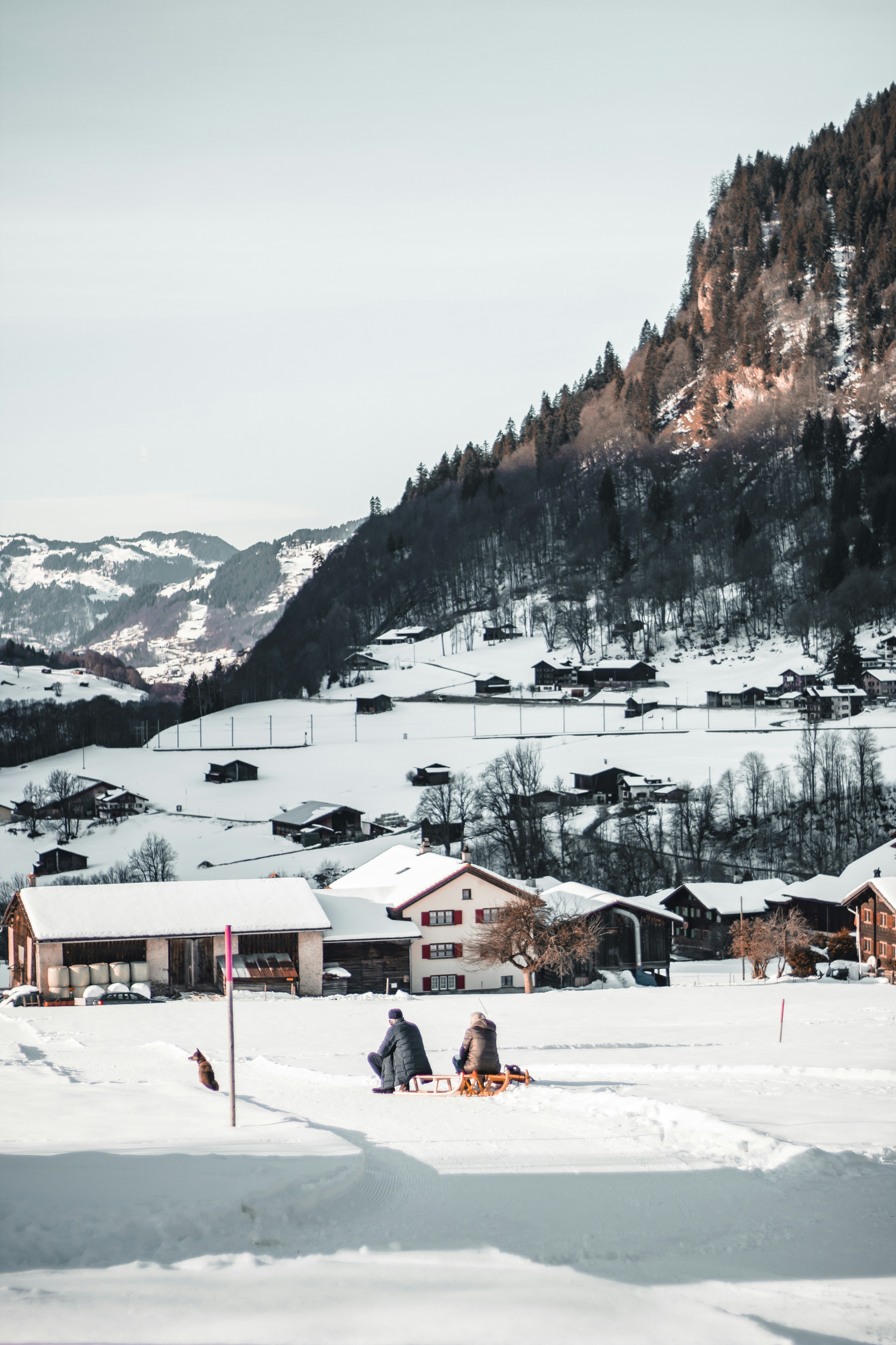 Two figures sit on sleds in a snowy landscape, surrounded by quaint houses and a towering mountain backdrop. The scene captures a serene winter day.