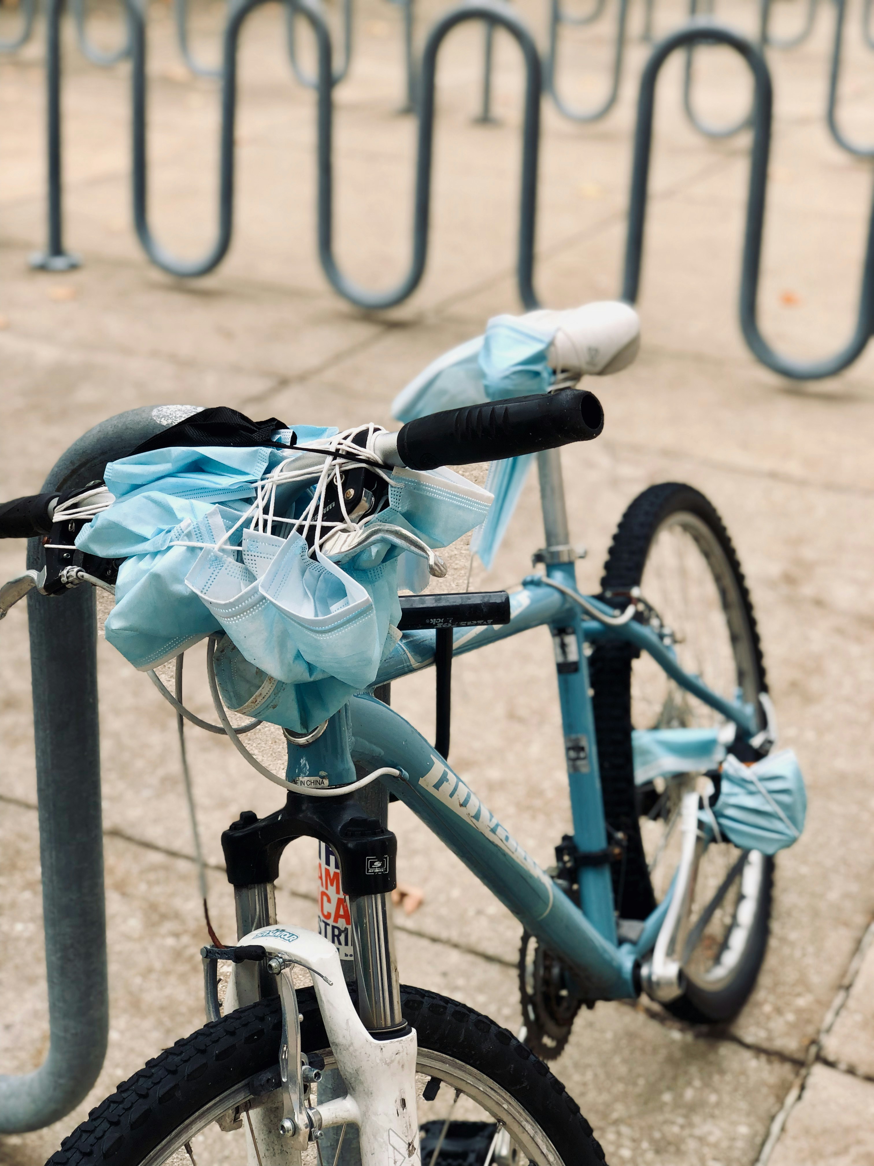 A blue bicycle adorned with light blue protective covers on the handlebars, parked in a bike rack. The setting showcases a blend of urban life and personal touches.