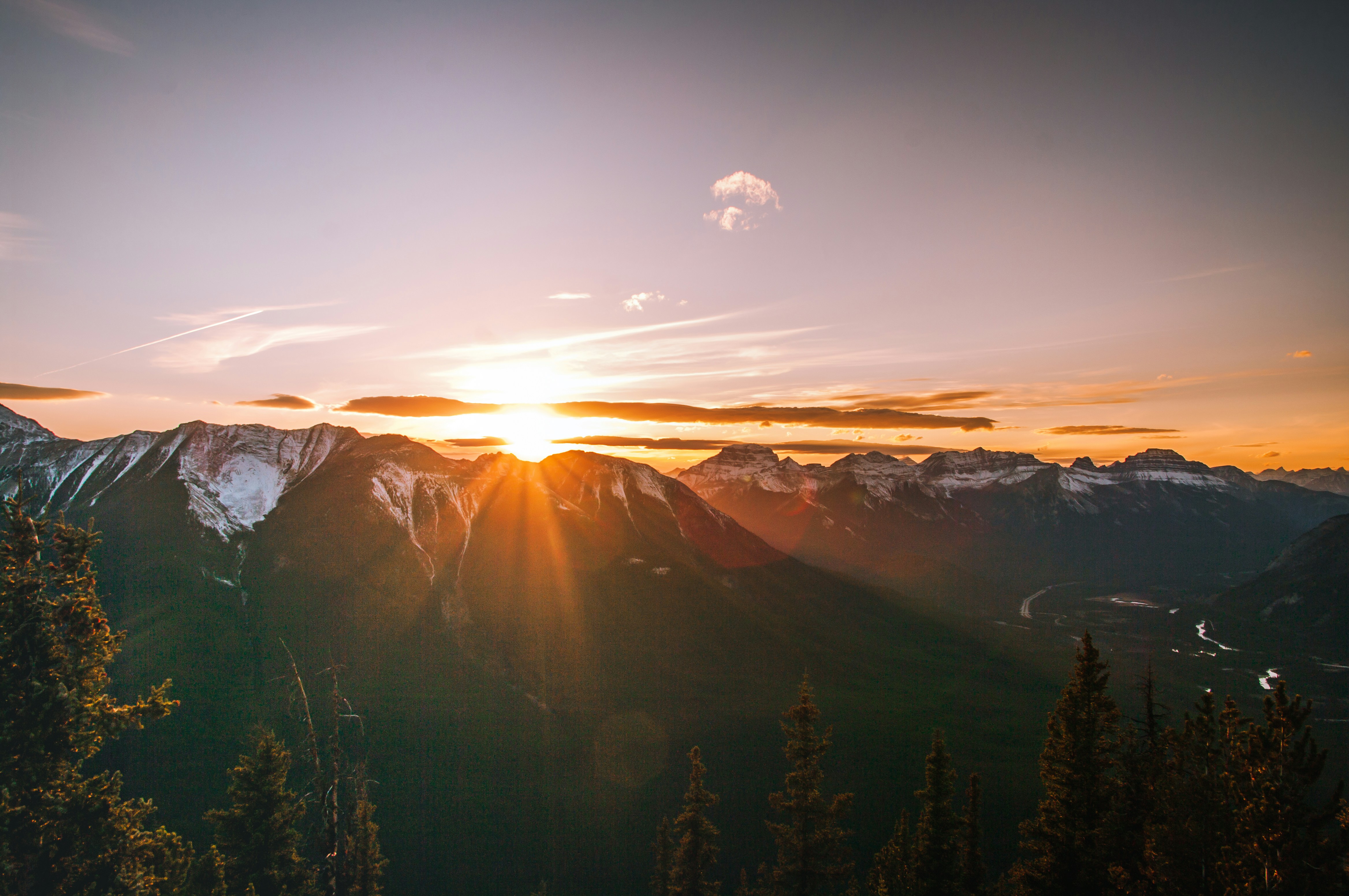 Sunrise over snow-capped peaks lights a pine-filled valley with warm, golden hues.