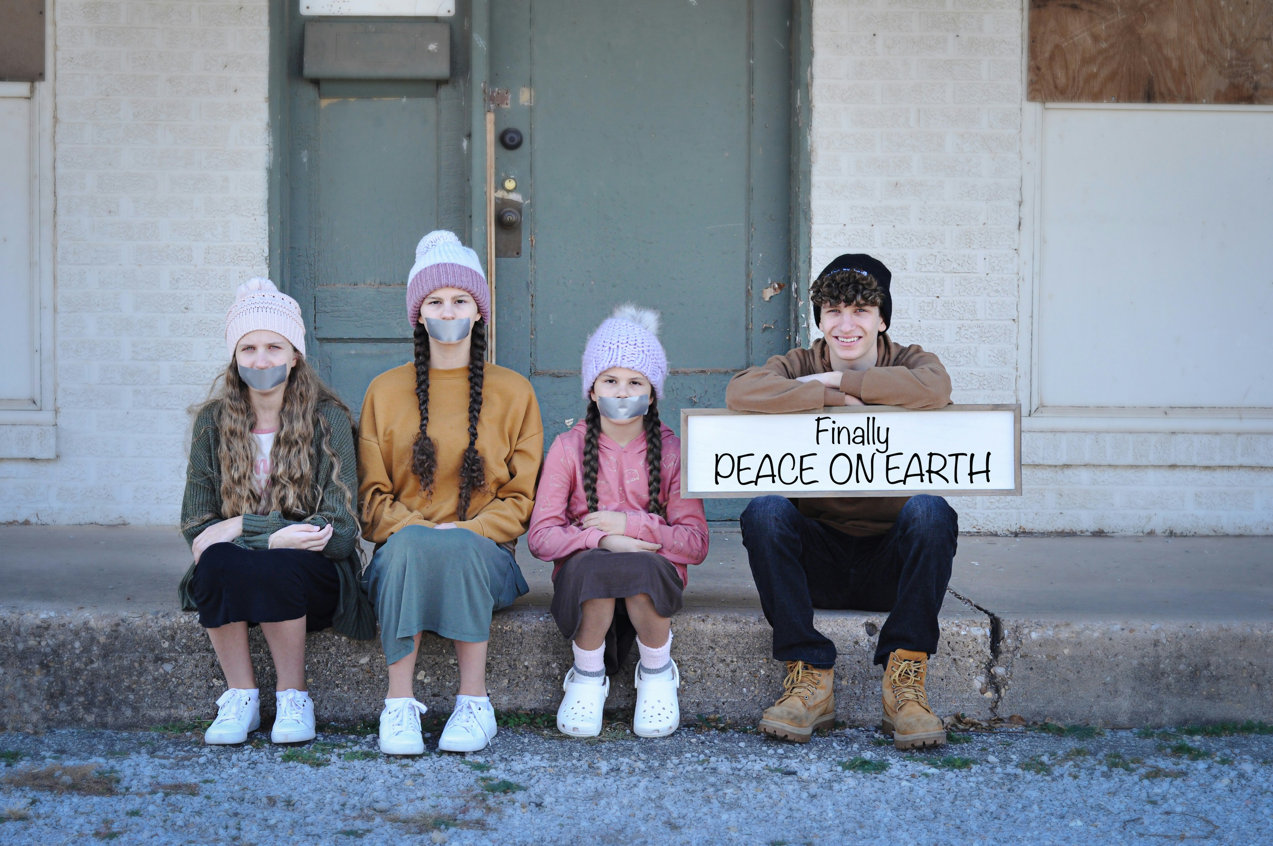 Four children sitting on steps, wearing winter hats and masks, holding a sign that reads 'Finally PEACE ON EARTH.'