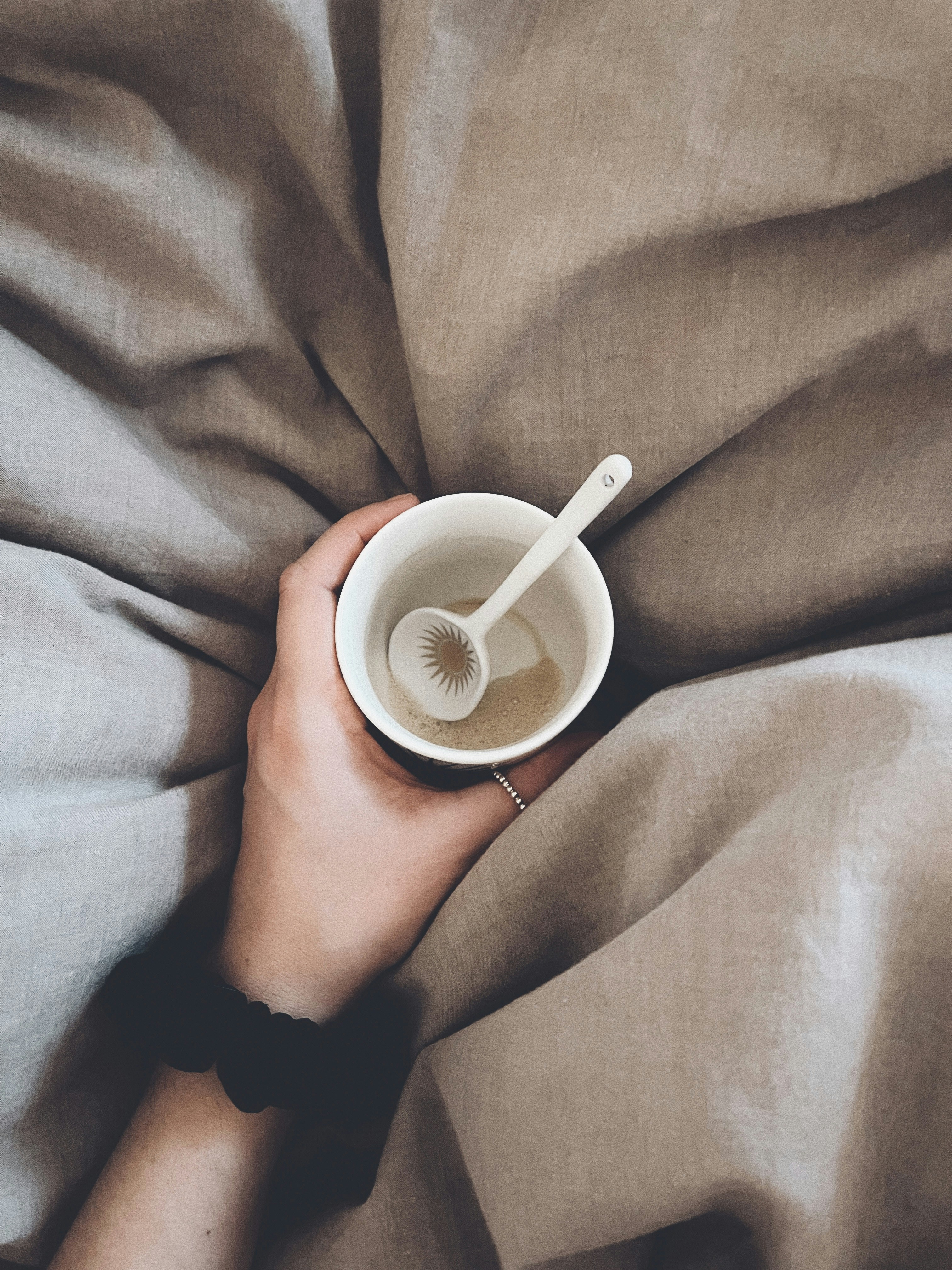 Hand holding a small cup with a spoon resting inside, surrounded by soft bedding textures.