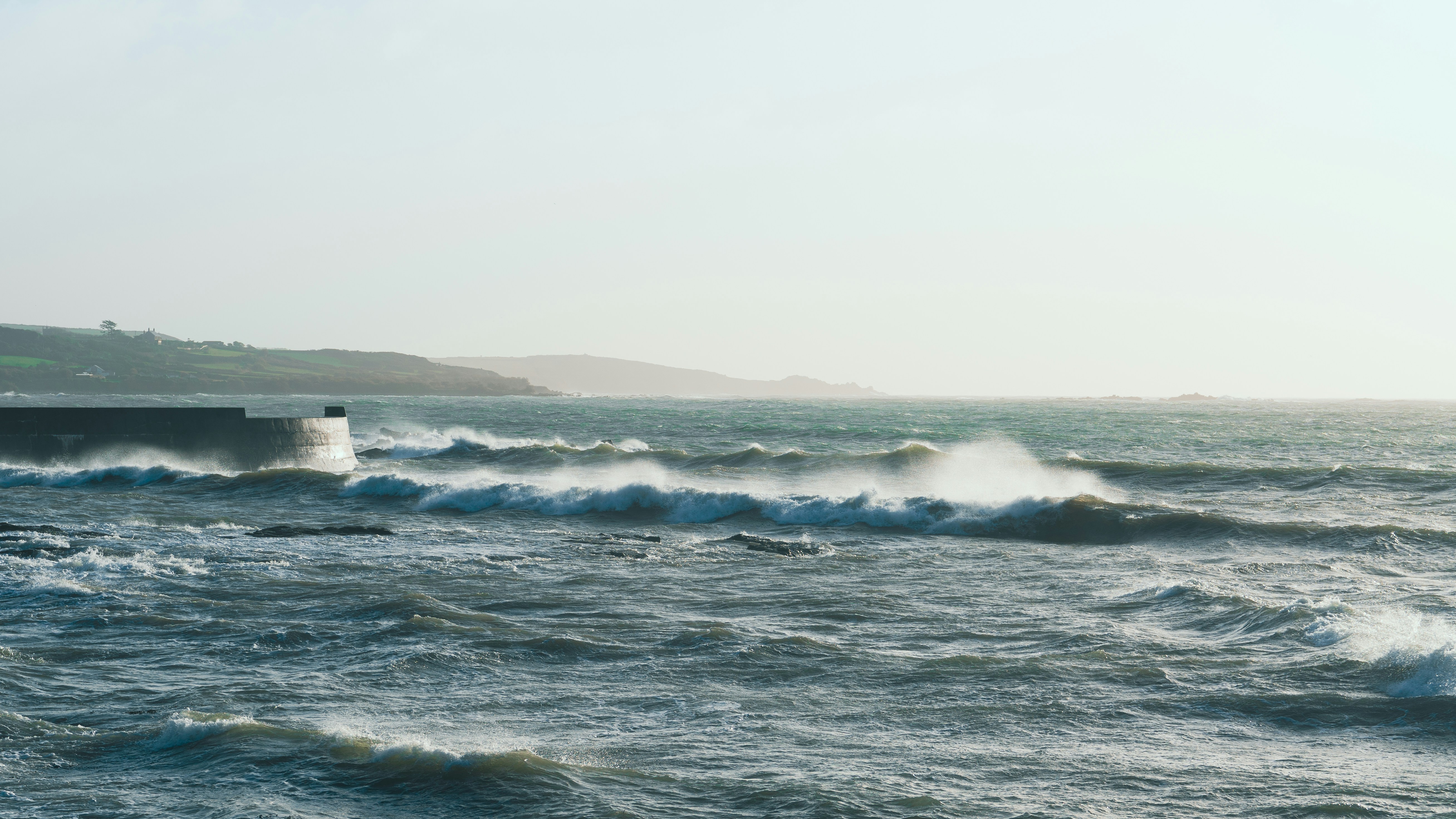Powerful ocean waves crashing against a coastal pier under a soft, hazy sky.