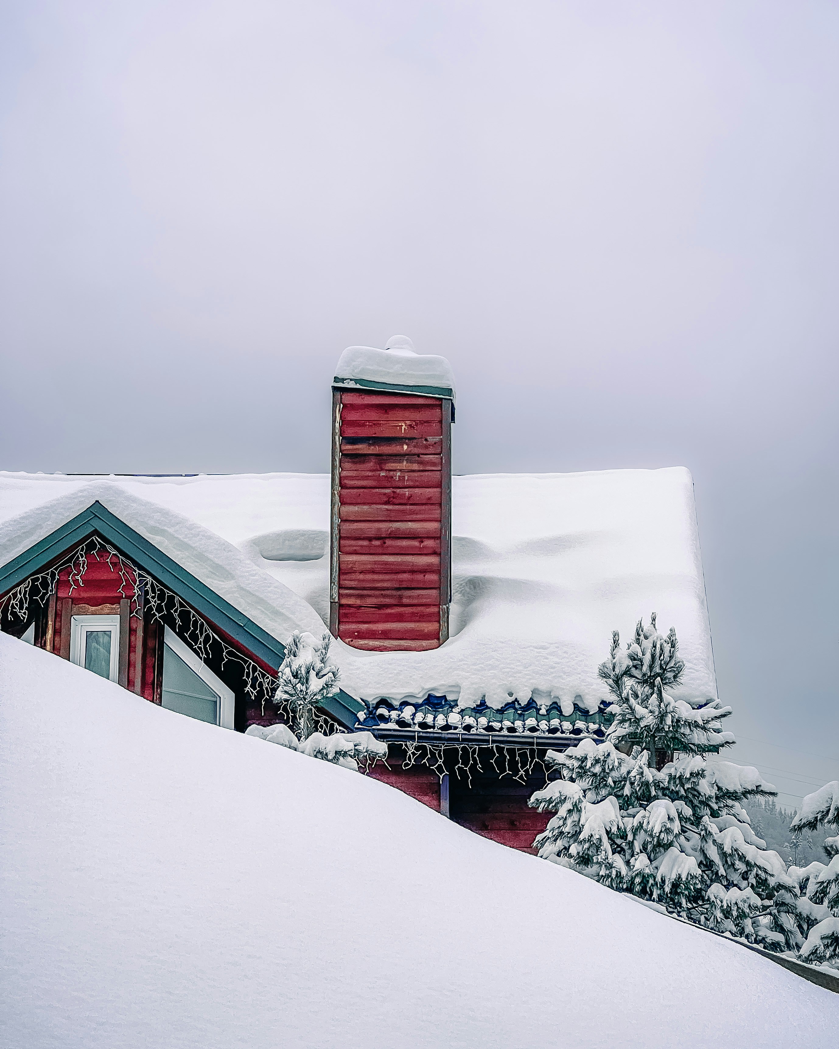 A charming red house partially buried under a blanket of snow, with icicles hanging from the eaves and a chimney peeking through the white landscape.