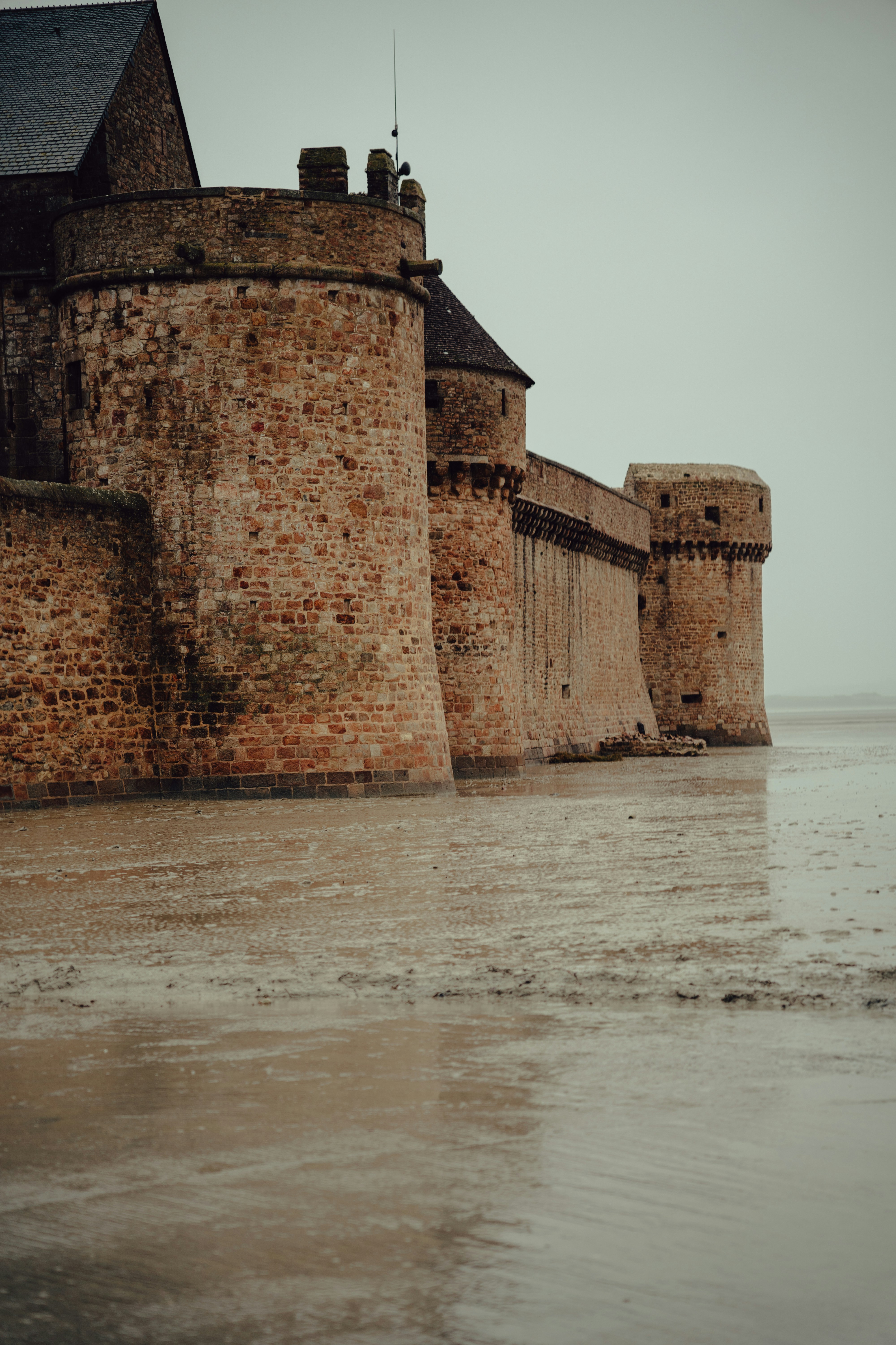 Ancient stone fortress standing resilient against the tide, surrounded by a reflective wet landscape under a moody sky.
