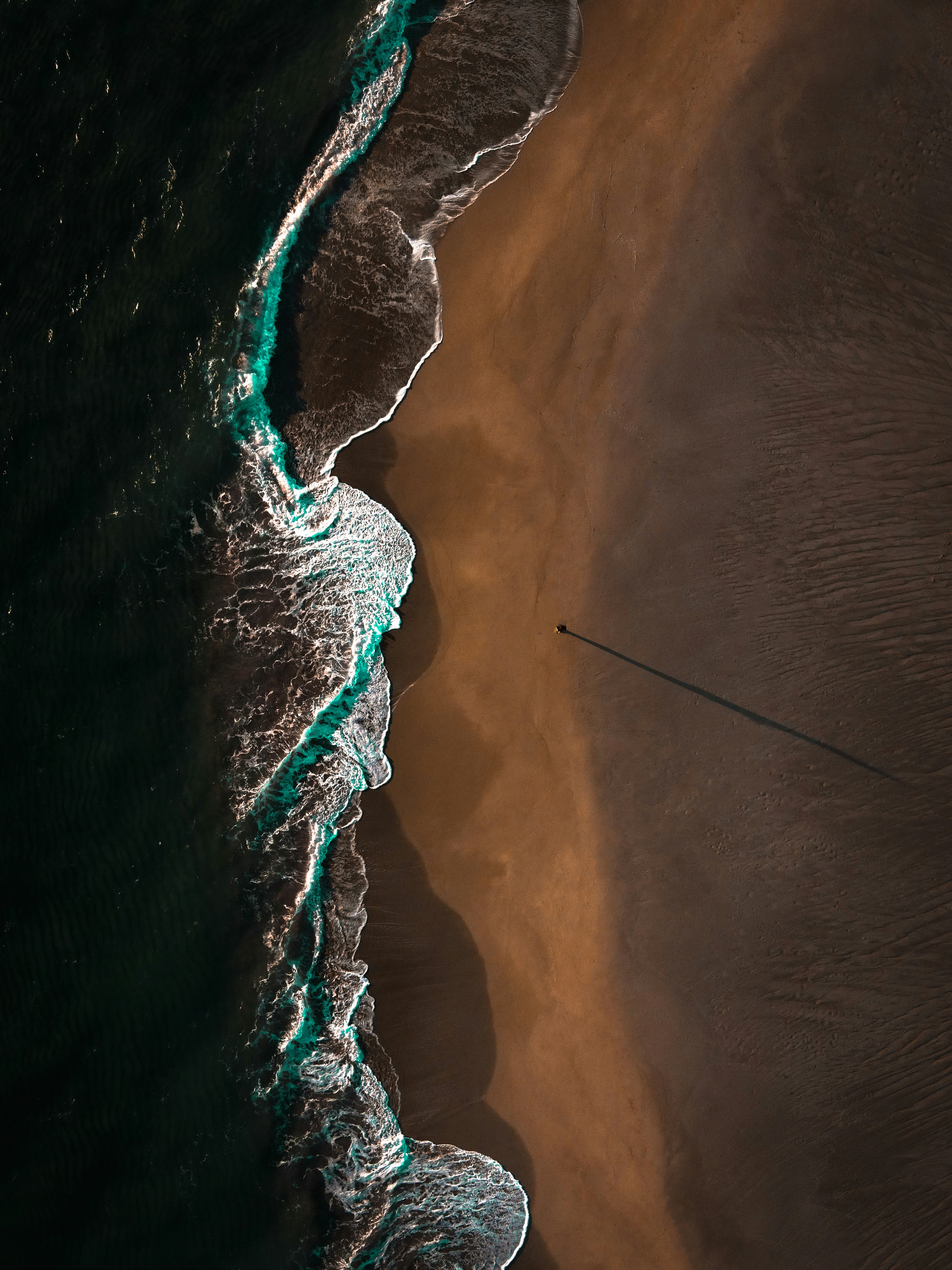 Aerial view of turquoise waves meeting a wide, sandy beach with a solitary person casting a long shadow.