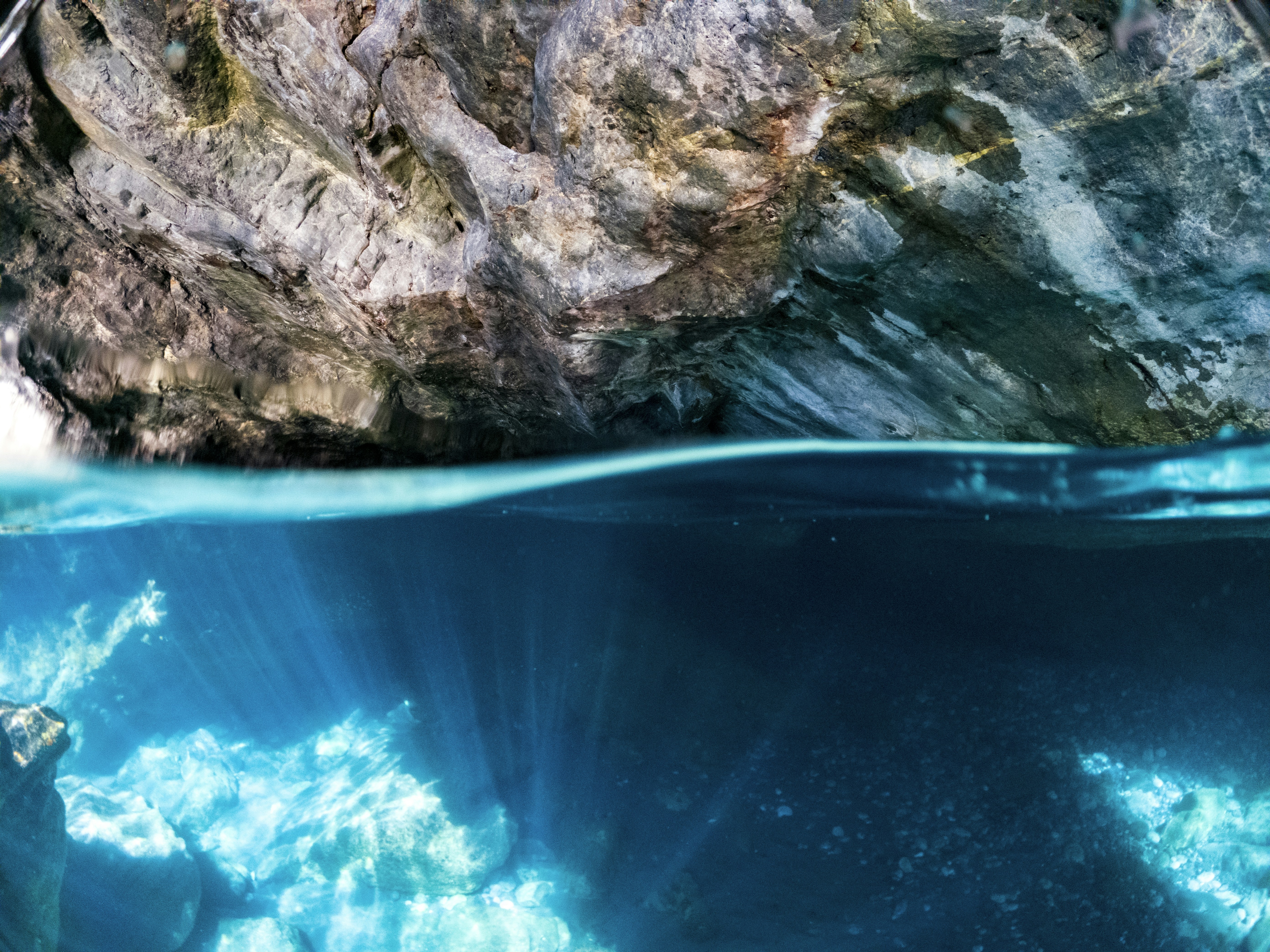 A half-submerged shot inside a rocky sea cave reveals sunlit blue water and rugged limestone above, with rays penetrating the clear depths. The scene emphasizes the boundary between air and water and the cave's textured walls.