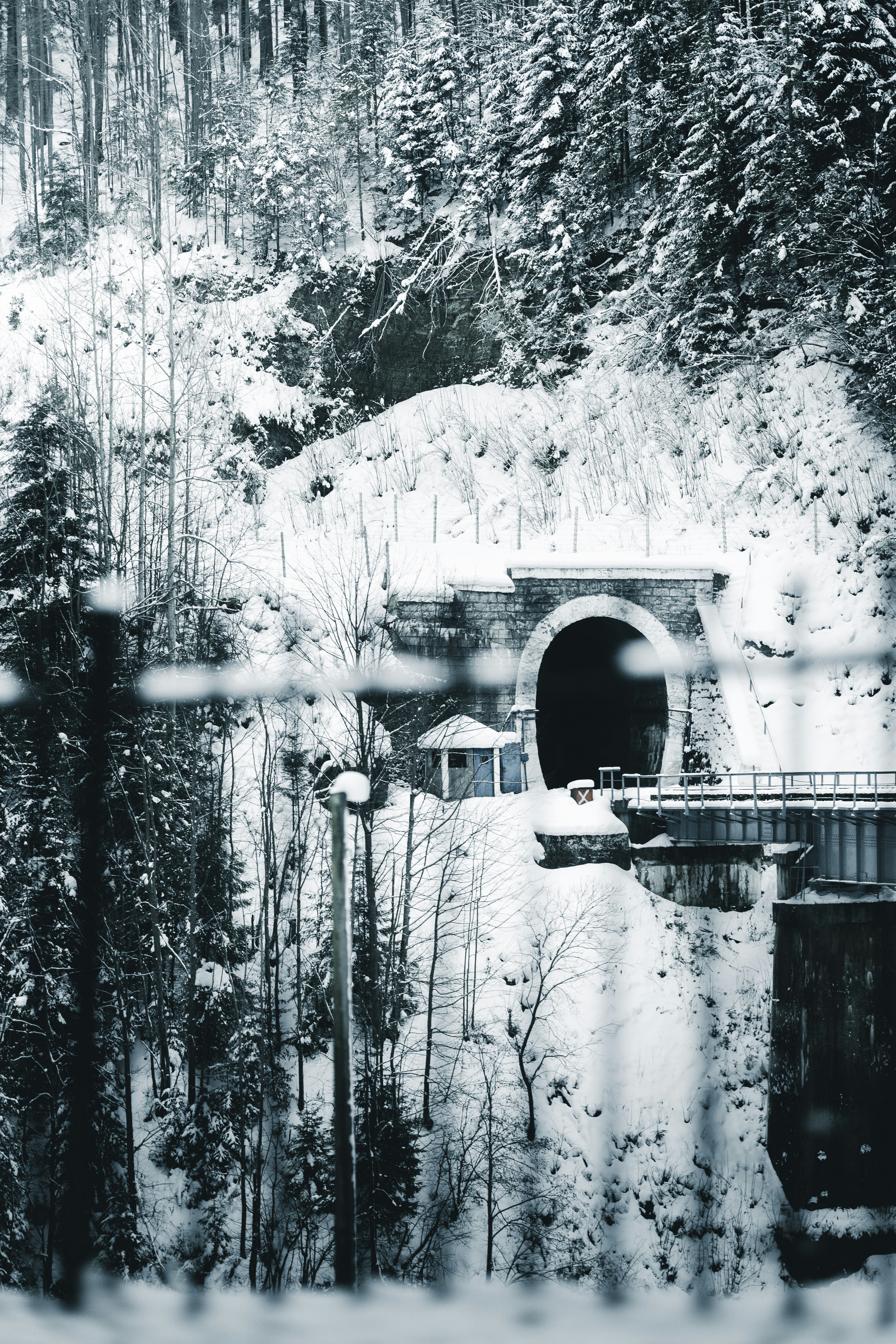 Snow-covered landscape revealing a dark tunnel nestled in the mountains, surrounded by frosted trees. A small structure stands nearby, hinting at human presence in this serene environment.