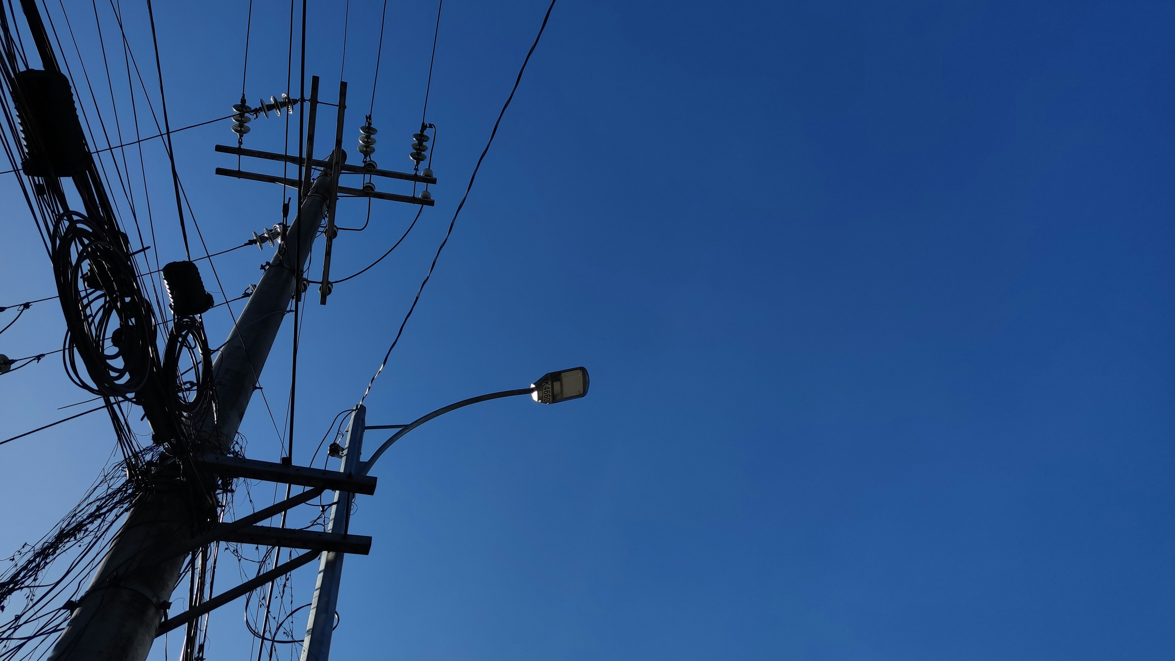 Electric utility pole adorned with tangled wires under a clear blue sky. A streetlight casts a soft glow.