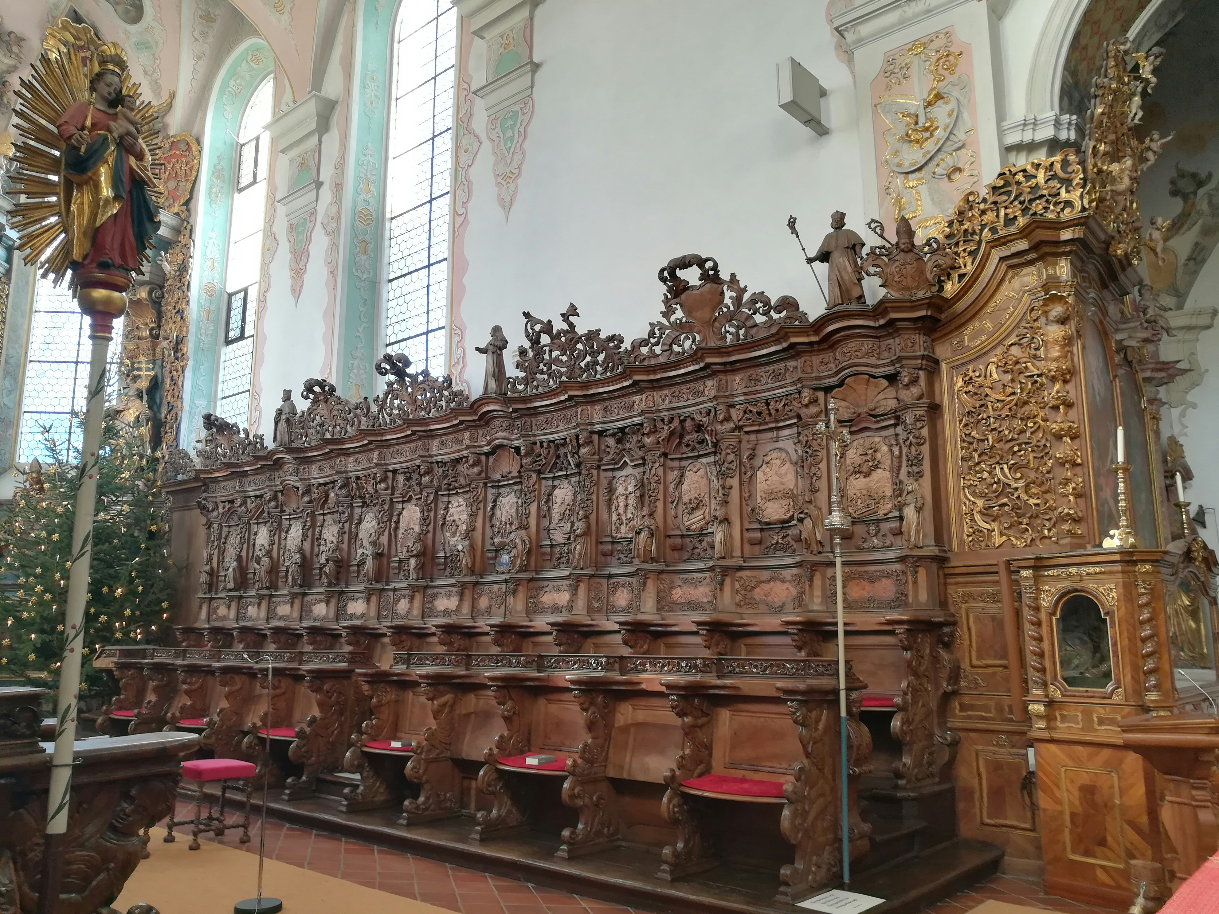 Intricately carved wooden choir stalls line a grand church interior, with tall arched windows and a glowing iconography above, inviting reverent contemplation.