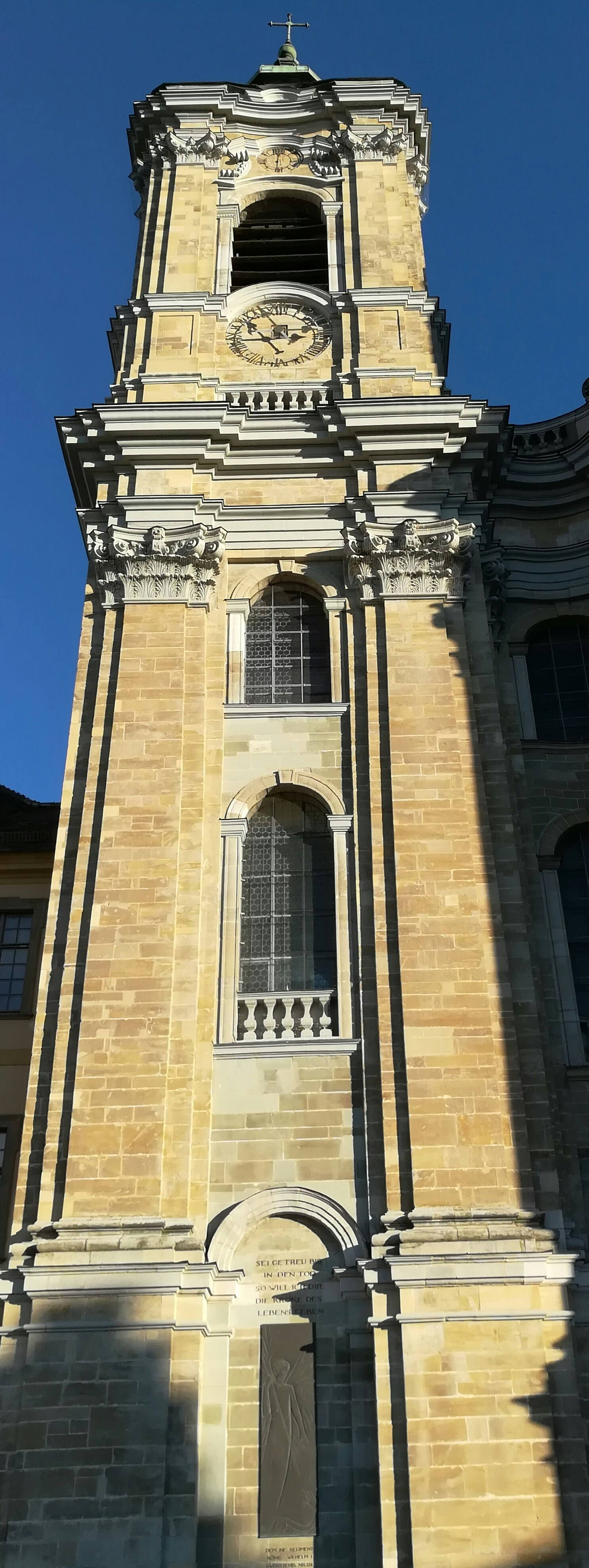 Historic clock tower rising against a clear blue sky, showcasing intricate architectural details and shadows. A bronze plaque adds a touch of artistry to the stone facade.