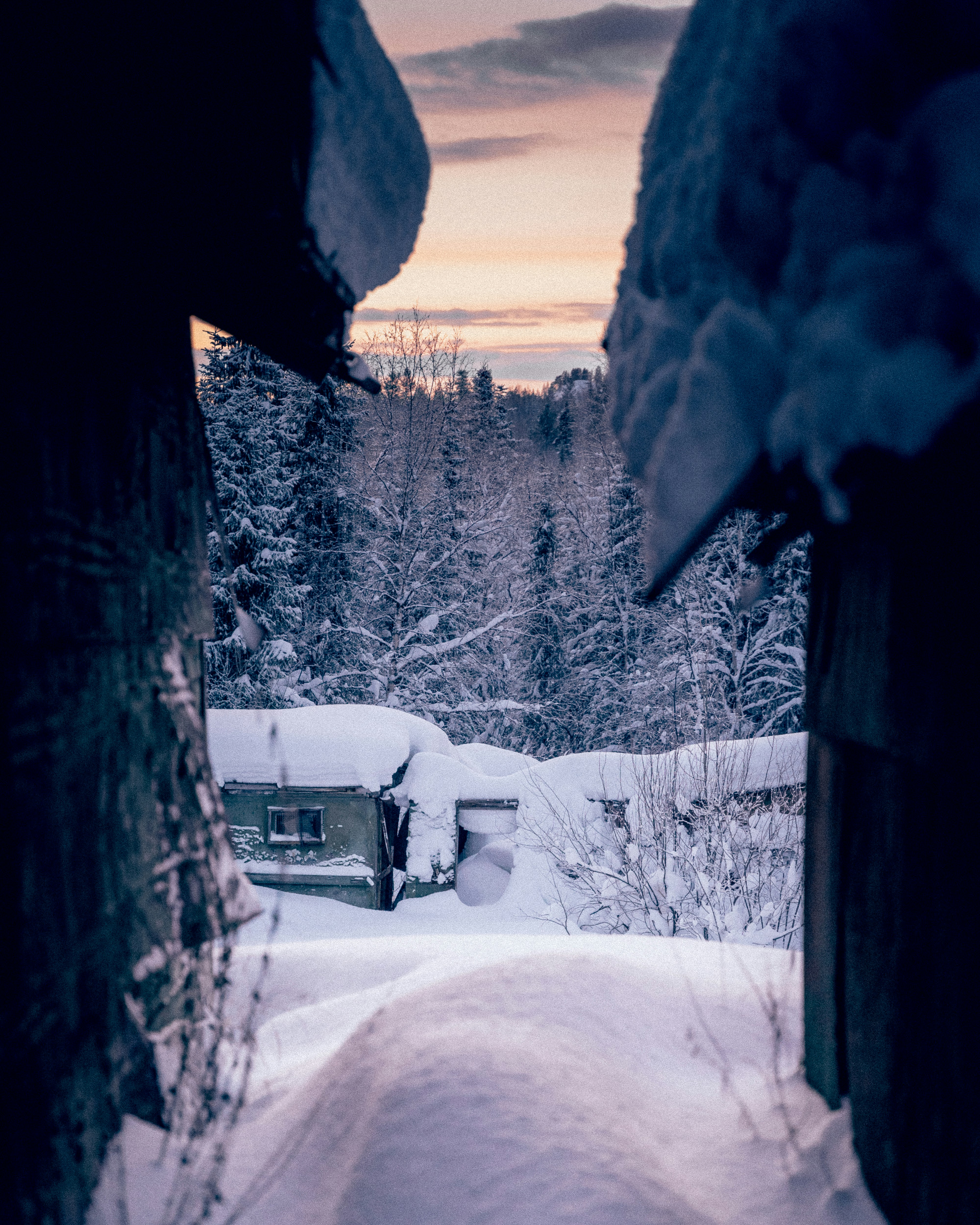 A serene winter scene framed by rustic wooden structures, revealing a snow-covered landscape and a hint of a vintage green cabin. The soft light of dusk casts a tranquil atmosphere.