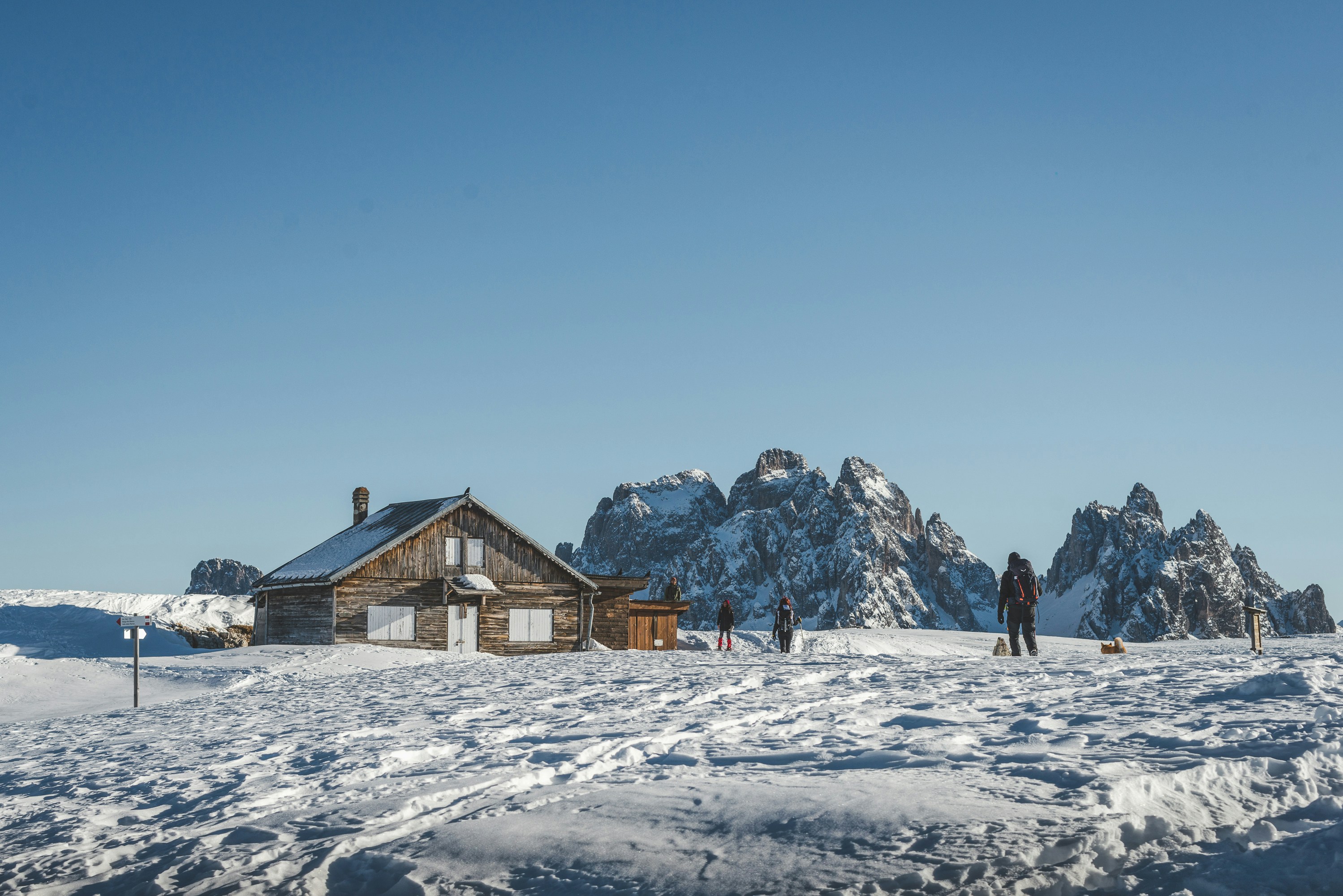 A rustic wooden cabin nestled in a snowy landscape, surrounded by towering mountain peaks under a clear blue sky.
