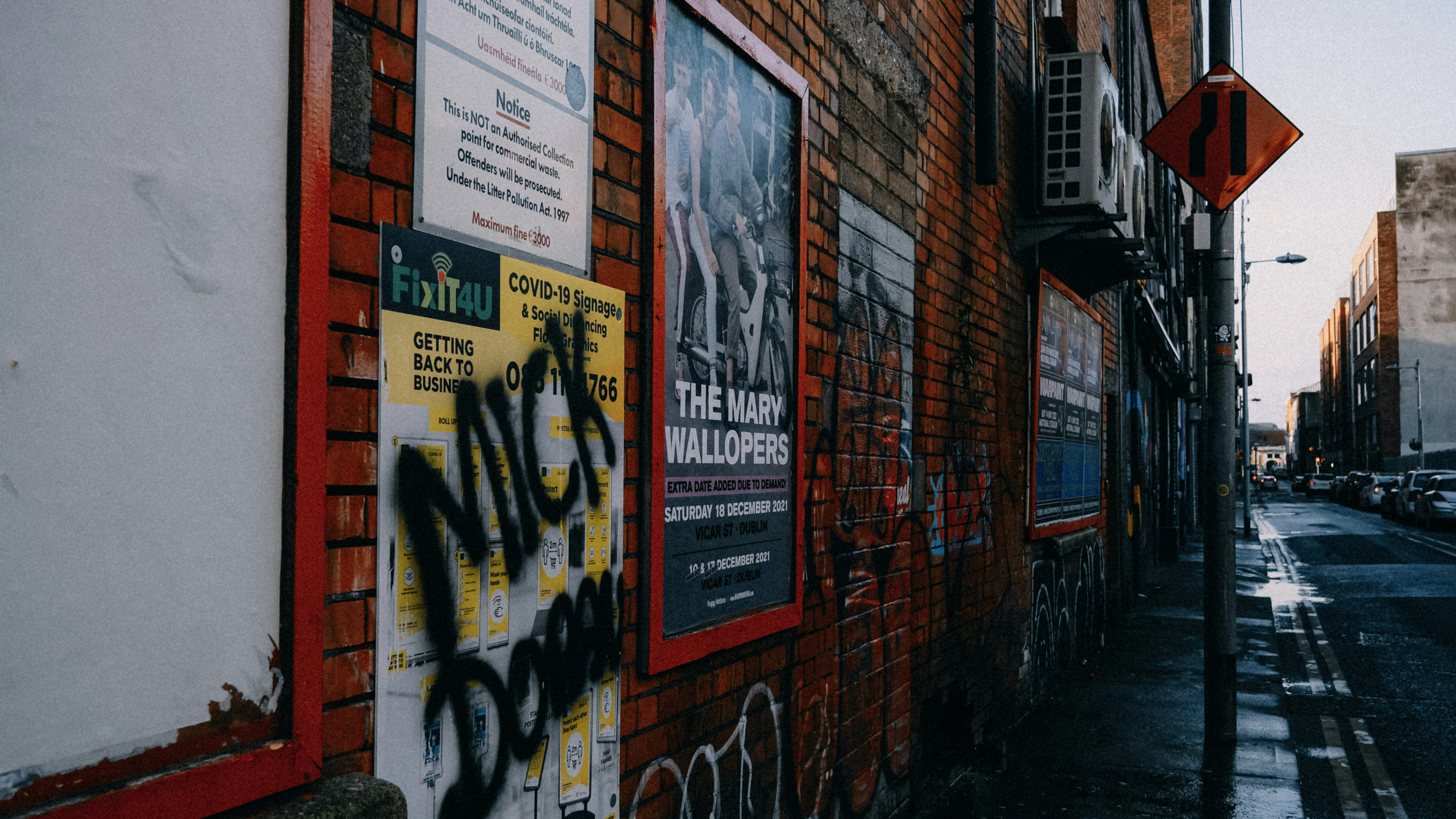 Graffiti and posters on a brick wall lining a narrow urban alleyway at dusk.