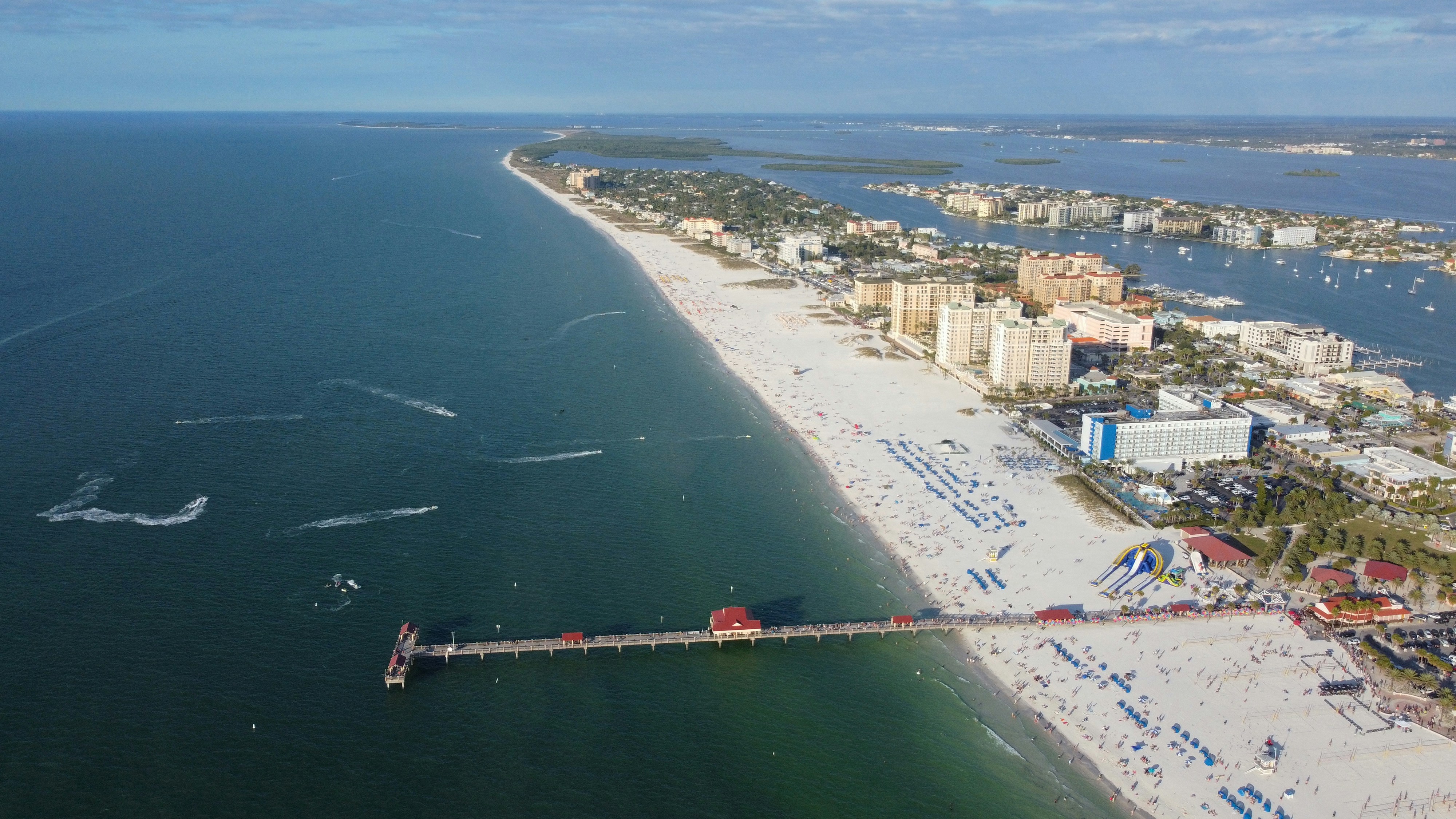 Aerial view of a long pier extending into the ocean alongside a bustling beachfront with buildings and boats in the distance.