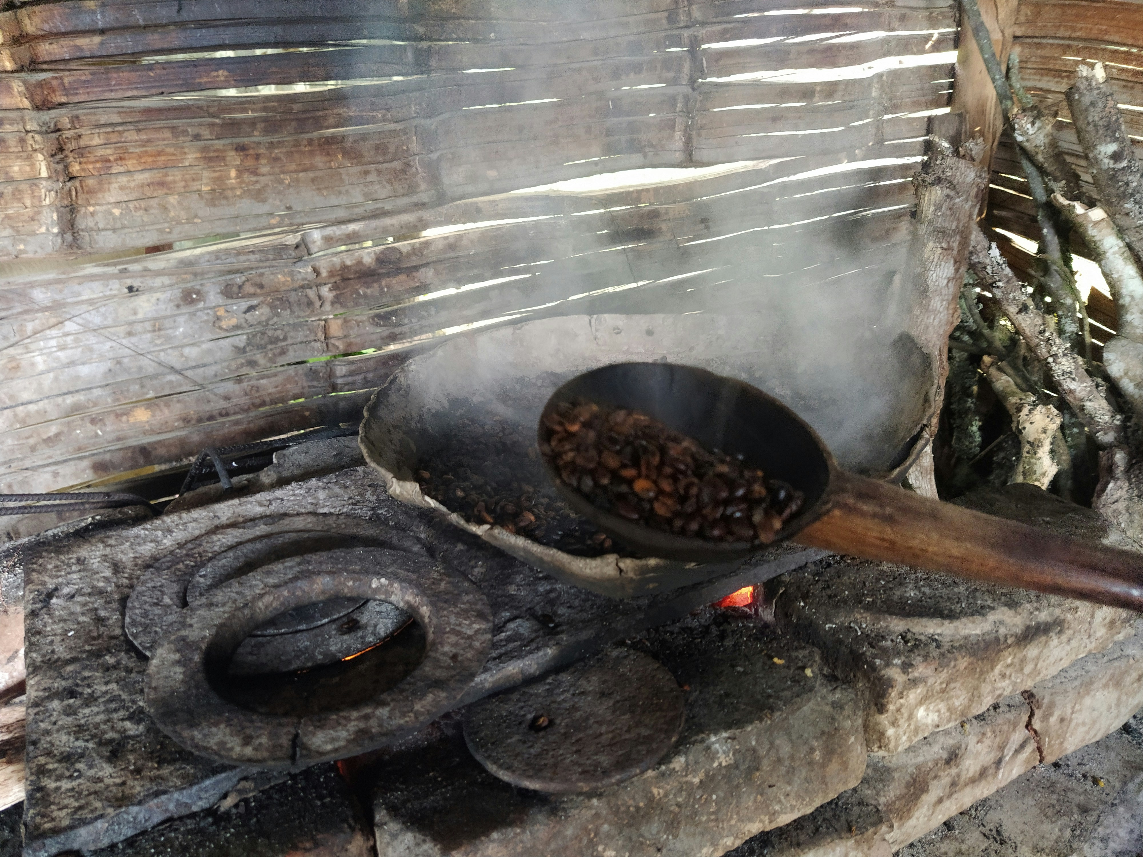 Wooden spoon holding roasted coffee beans over a smoky fire in a rustic kitchen. The scene captures the traditional coffee roasting process.