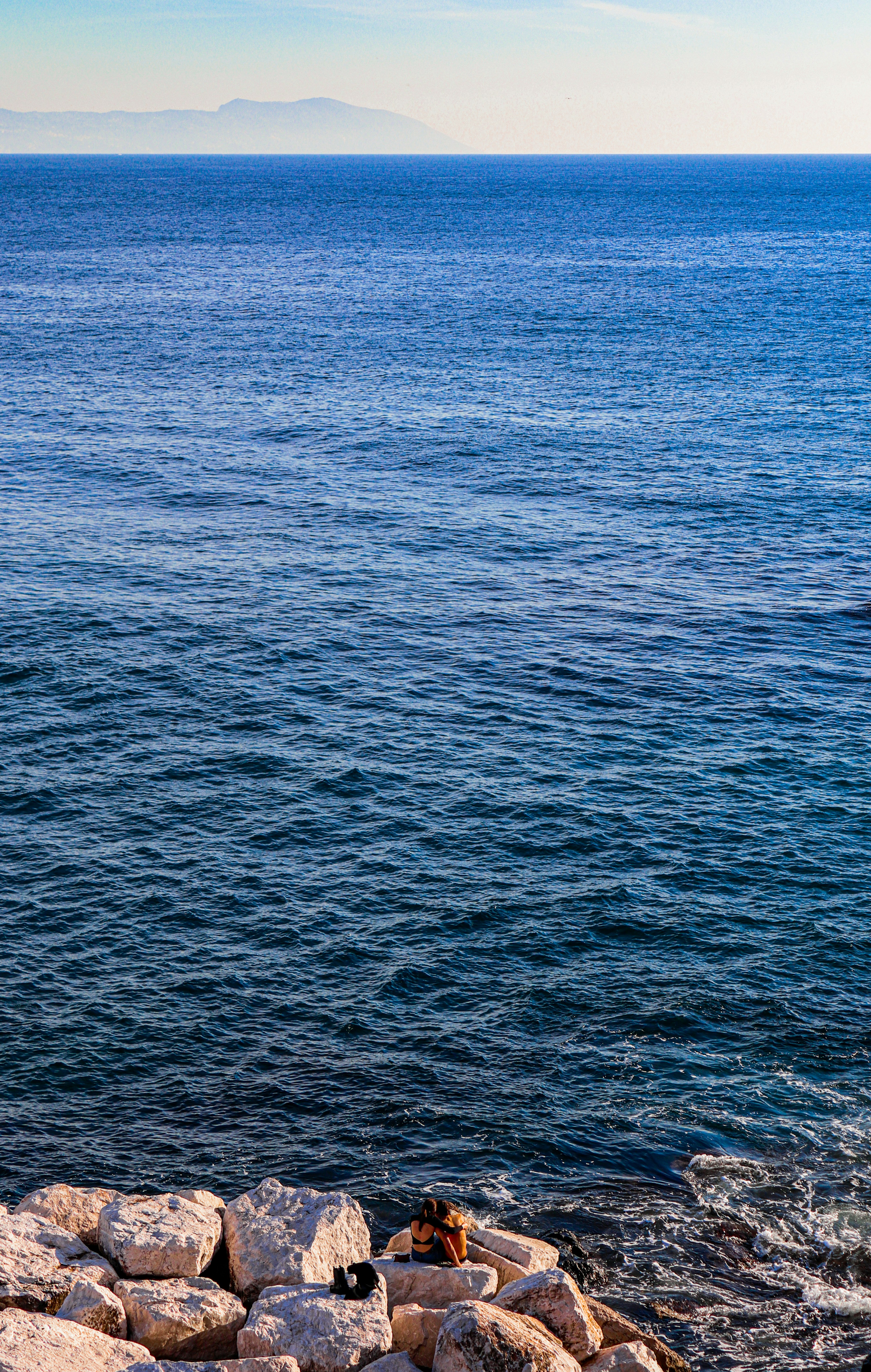 A person sitting on a rocky outcrop, gazing out at the calm sea under a clear sky. The tranquil waters reflect the soft hues of the horizon.