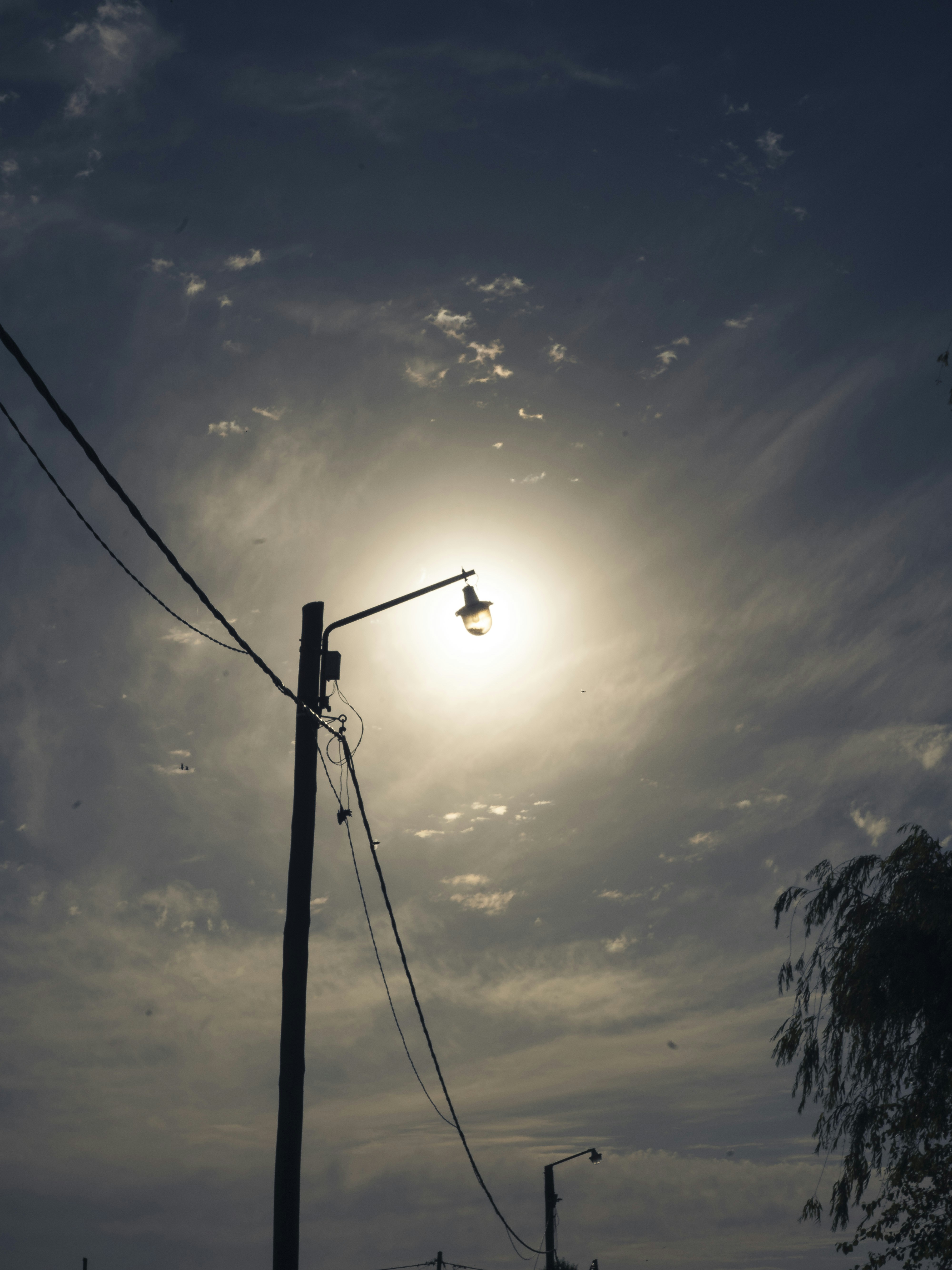 A street lamp stands silhouetted against a radiant sun surrounded by wispy clouds. The interplay of light creates a dramatic atmosphere.