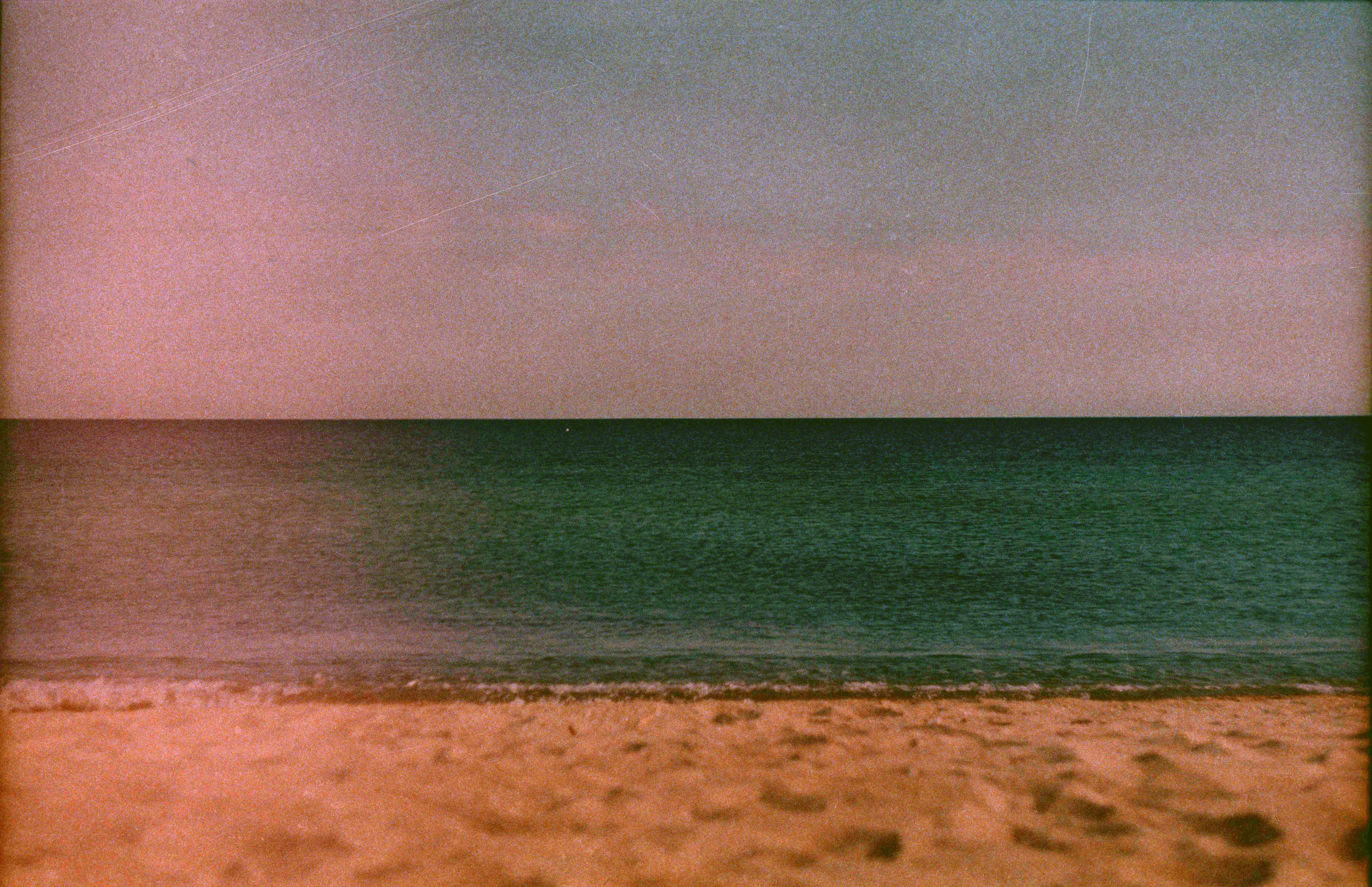 Film-scanned beach photograph showing a calm turquoise sea meeting a pale sky, with warm sand in the foreground. A nostalgic light-leak tint adds vintage mood.