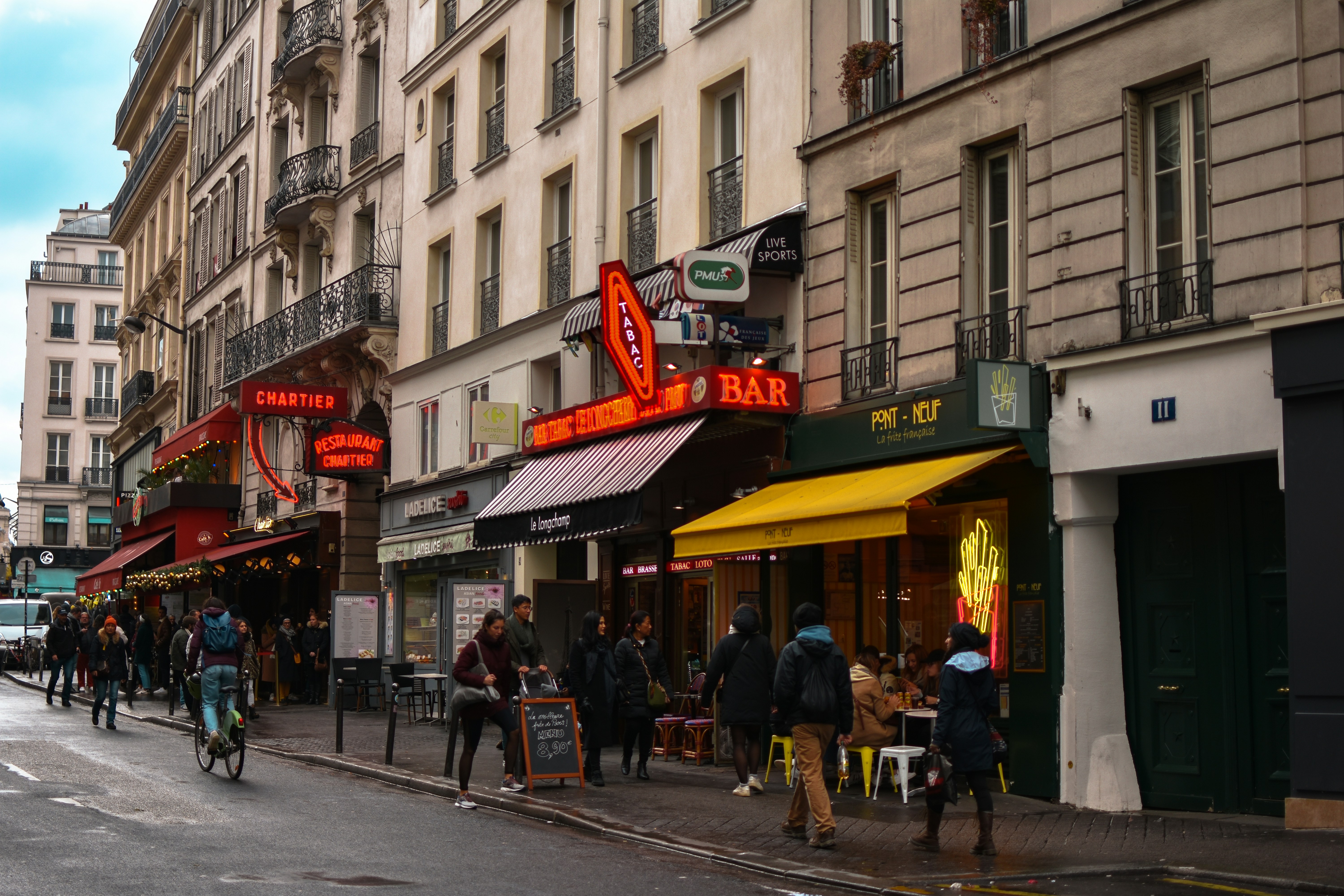 Bustling Parisian street featuring lively cafes and neon signs, inviting passersby to explore local cuisine and culture.