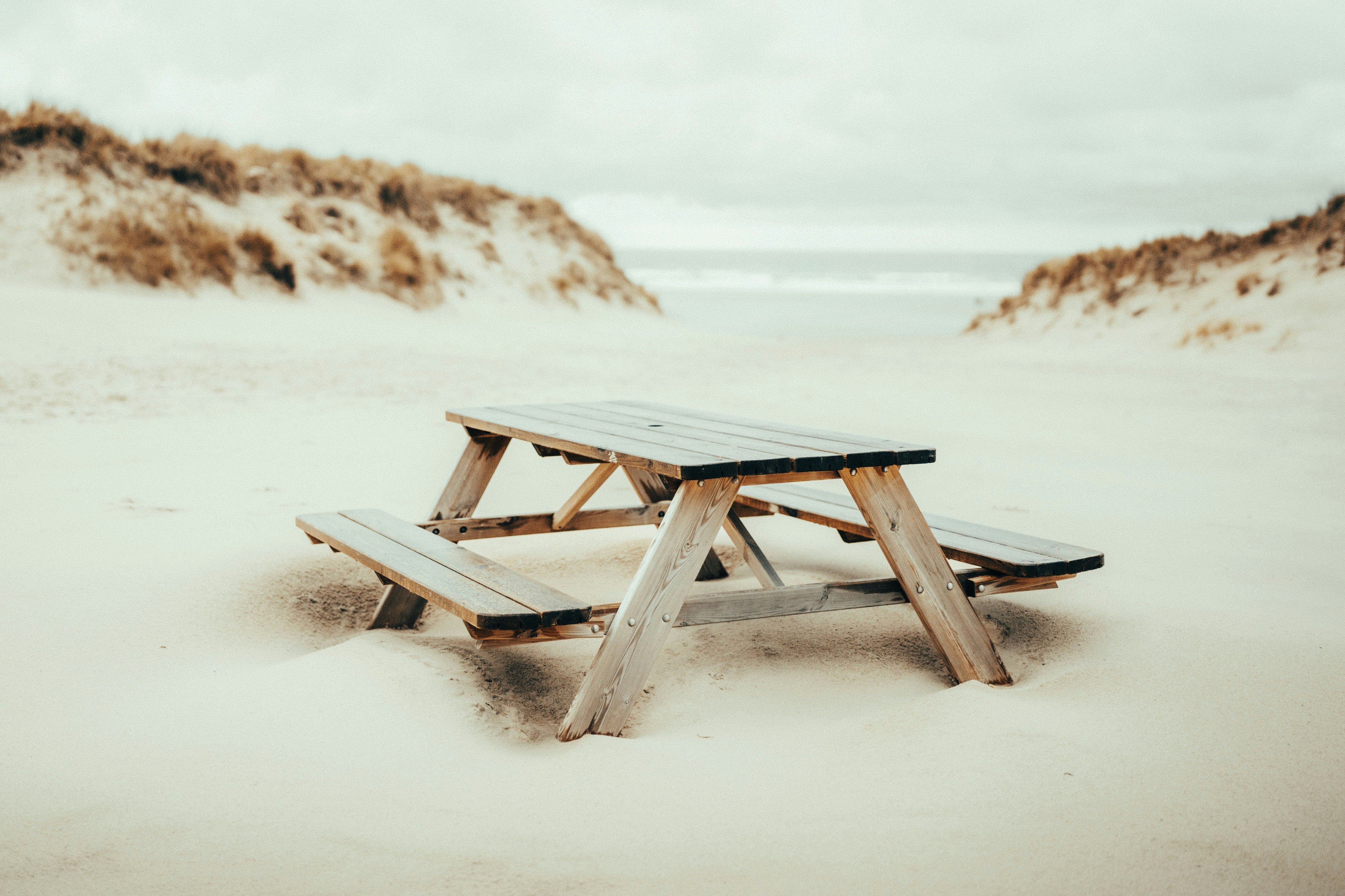 A weathered picnic table stands alone on a sandy beach, surrounded by gentle dunes and a muted sky. The scene evokes a sense of isolation and tranquility.